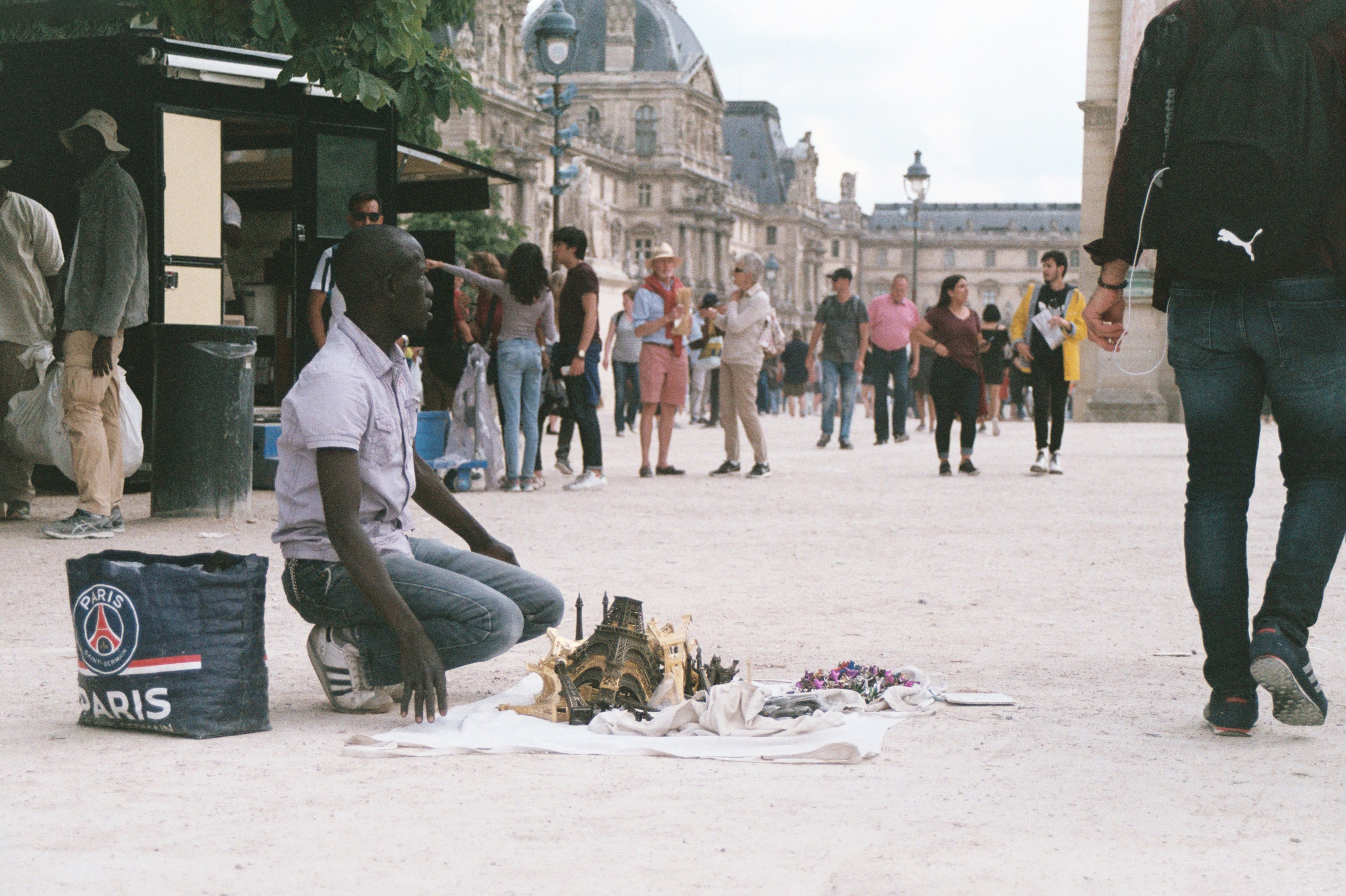 Photograph of a street vendor seated on the ground with pottery arranged on a cloth, while pedestrians walk through a sunlit Paris square.