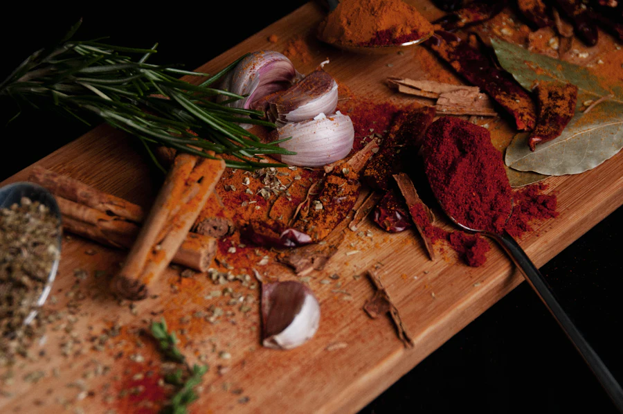Assorted spices, garlic cloves, cinnamon sticks, and herbs scattered across a wooden cutting board with a spoon of red seasoning. Dark background emphasizes the warm, rustic kitchen setup.