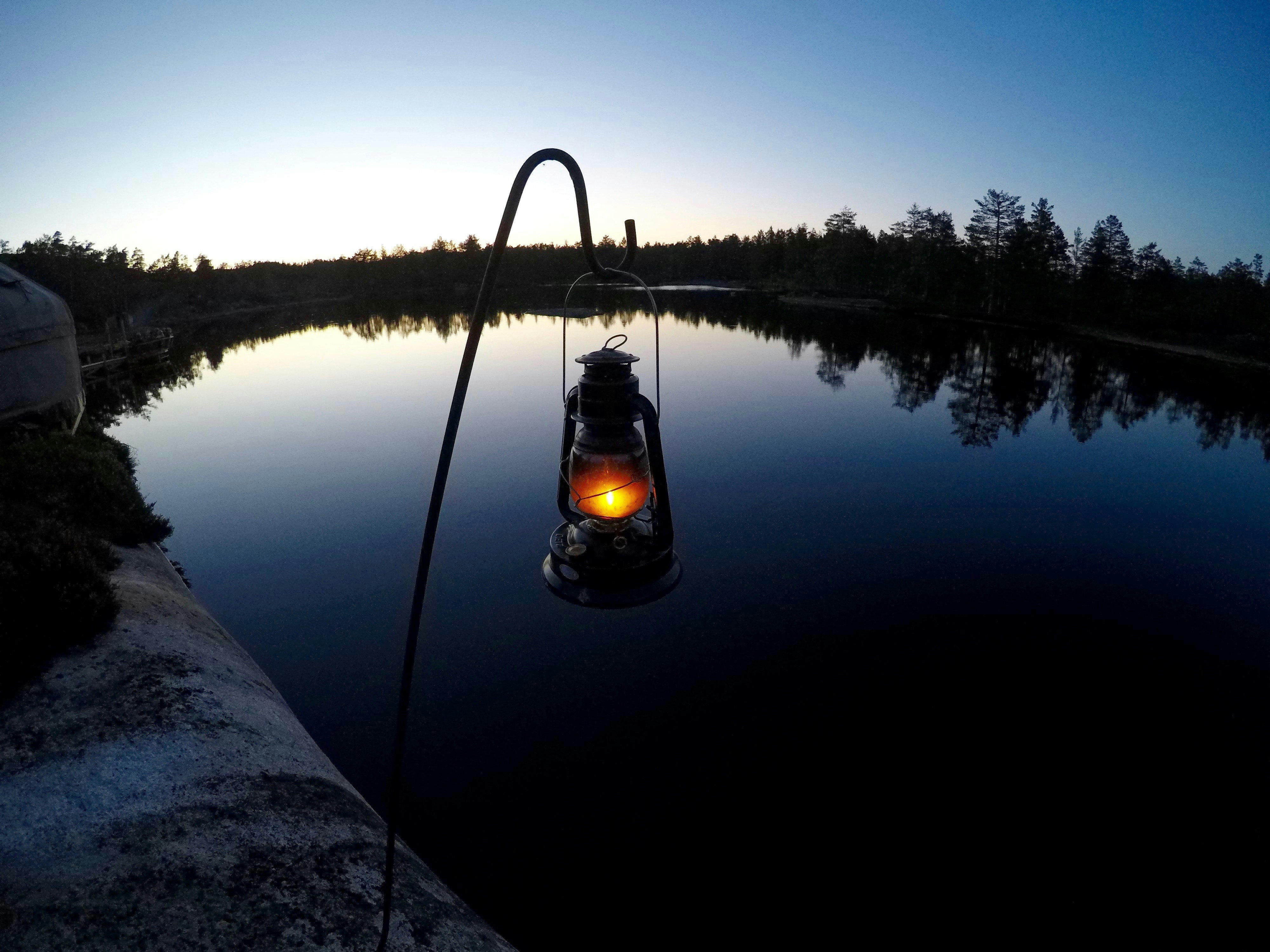 A vintage lantern hangs over a tranquil lake, its warm glow contrasting with the cool twilight sky. The still water mirrors the serene scene.