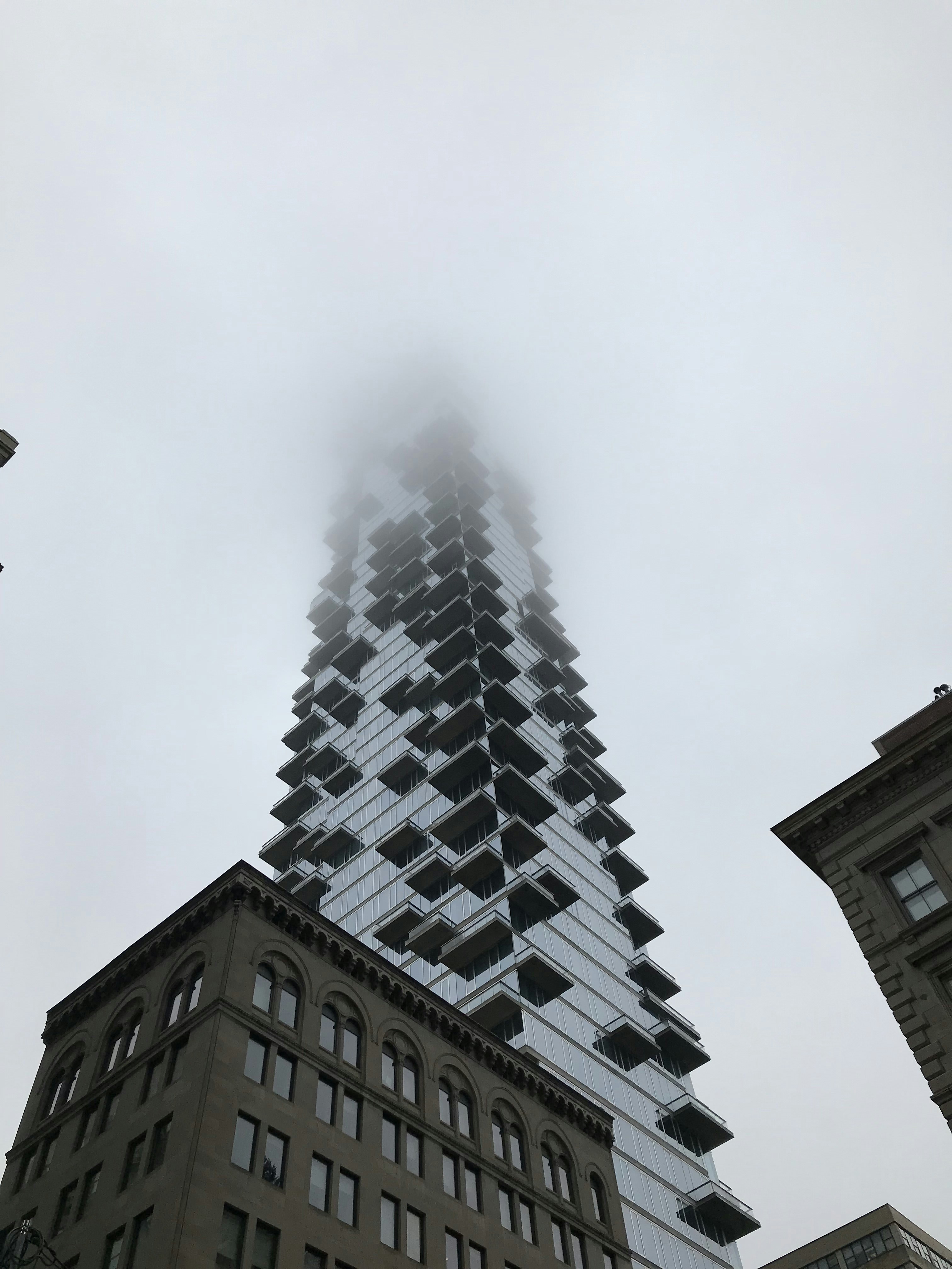 A modern skyscraper disappearing into thick fog, surrounded by historic buildings. The contrast between old and new architecture creates a striking urban scene.