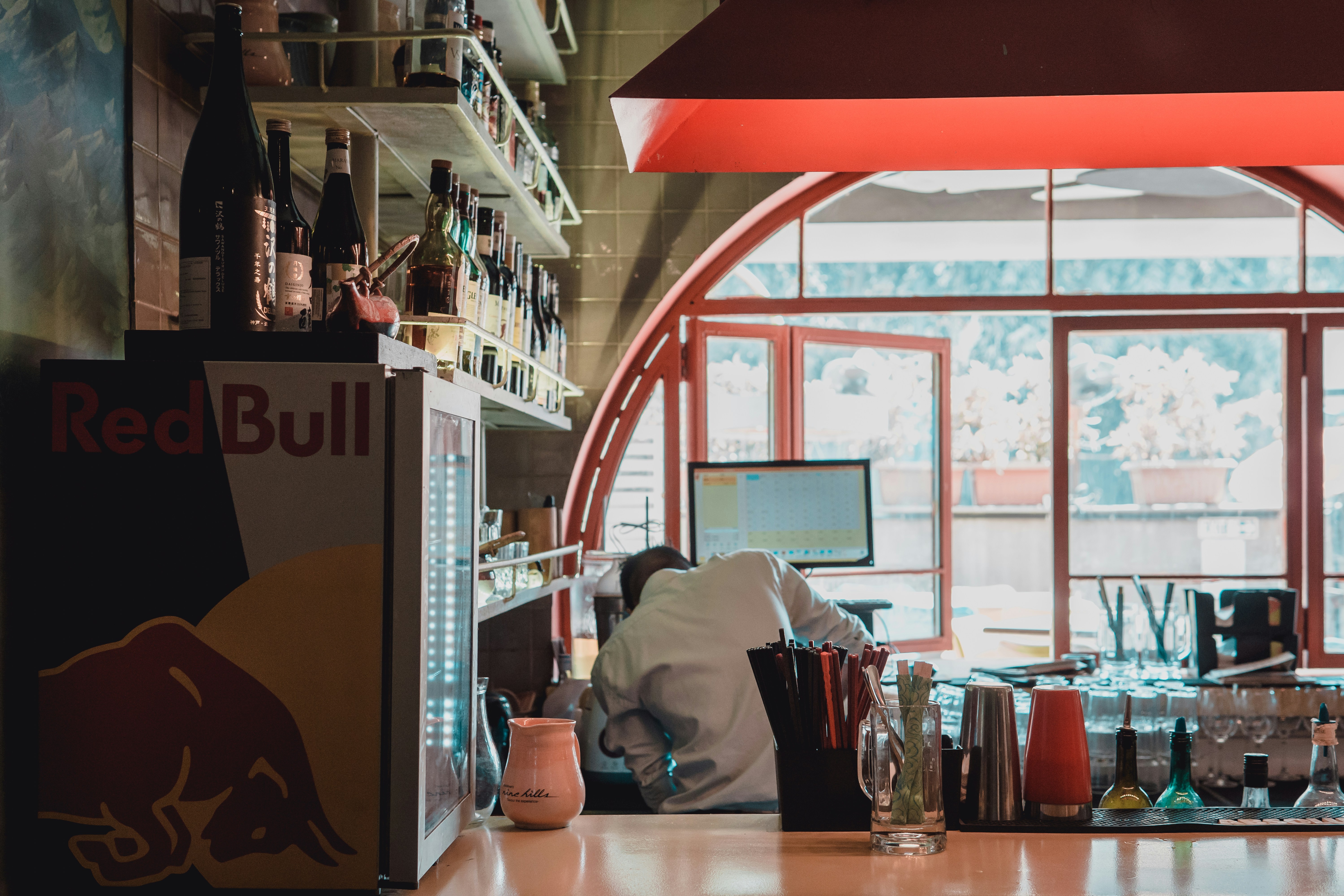Bartender organizing drinks in a vibrant bar setting with a Red Bull cooler and colorful bottles on the shelf.