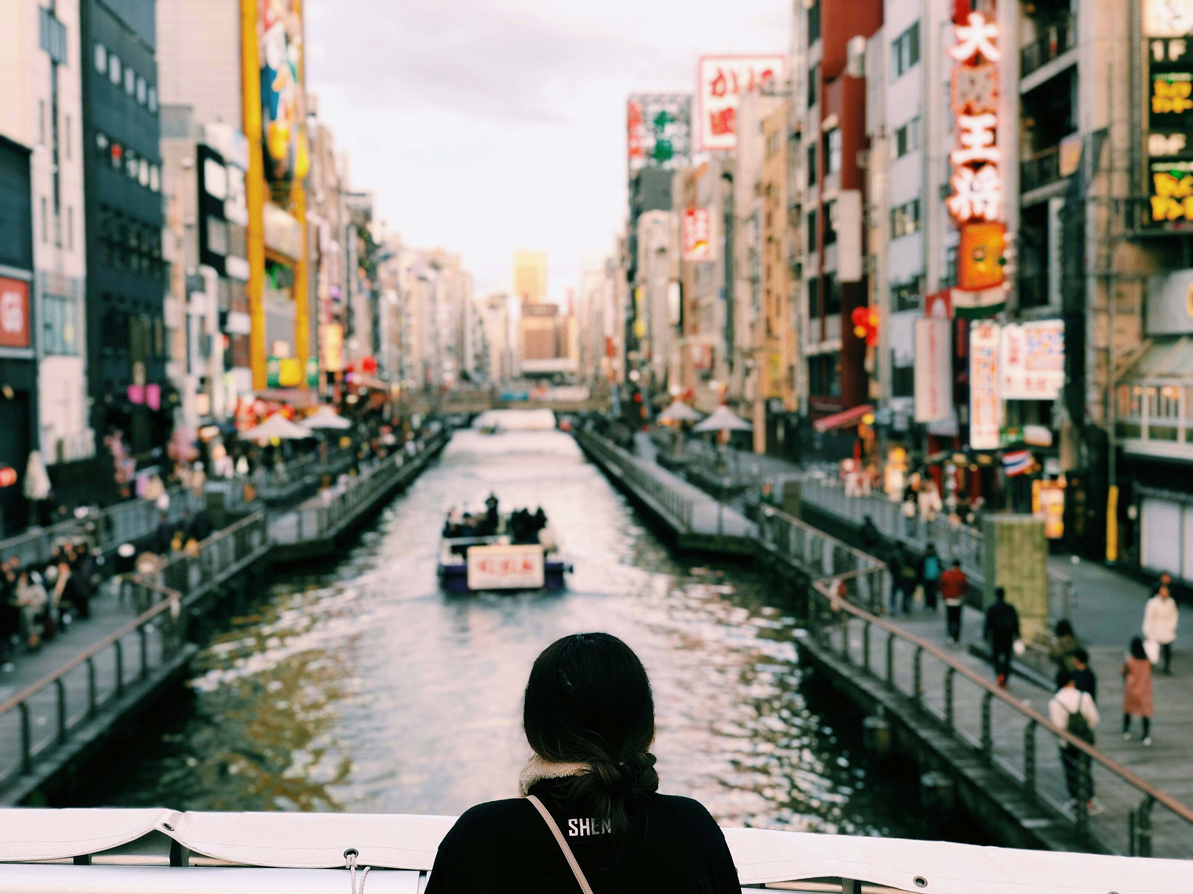 The image captures a tranquil moment in a bustling cityscape, where a solitary figure gazes over a vibrant canal flanked by lively streets. The composition draws the eye along the waterway, framed by colorful buildings adorned with neon signs under a soft, diffused light. The harmonious blend of urban energy and serene reflection makes this scene visually striking, with a palette of muted tones contrasted by bright accents.