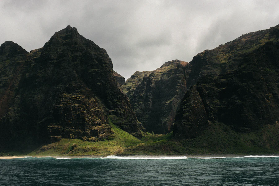 Steep dark cliffs rise above a narrow strip of green shoreline and choppy ocean water, with heavy clouds hanging overhead.