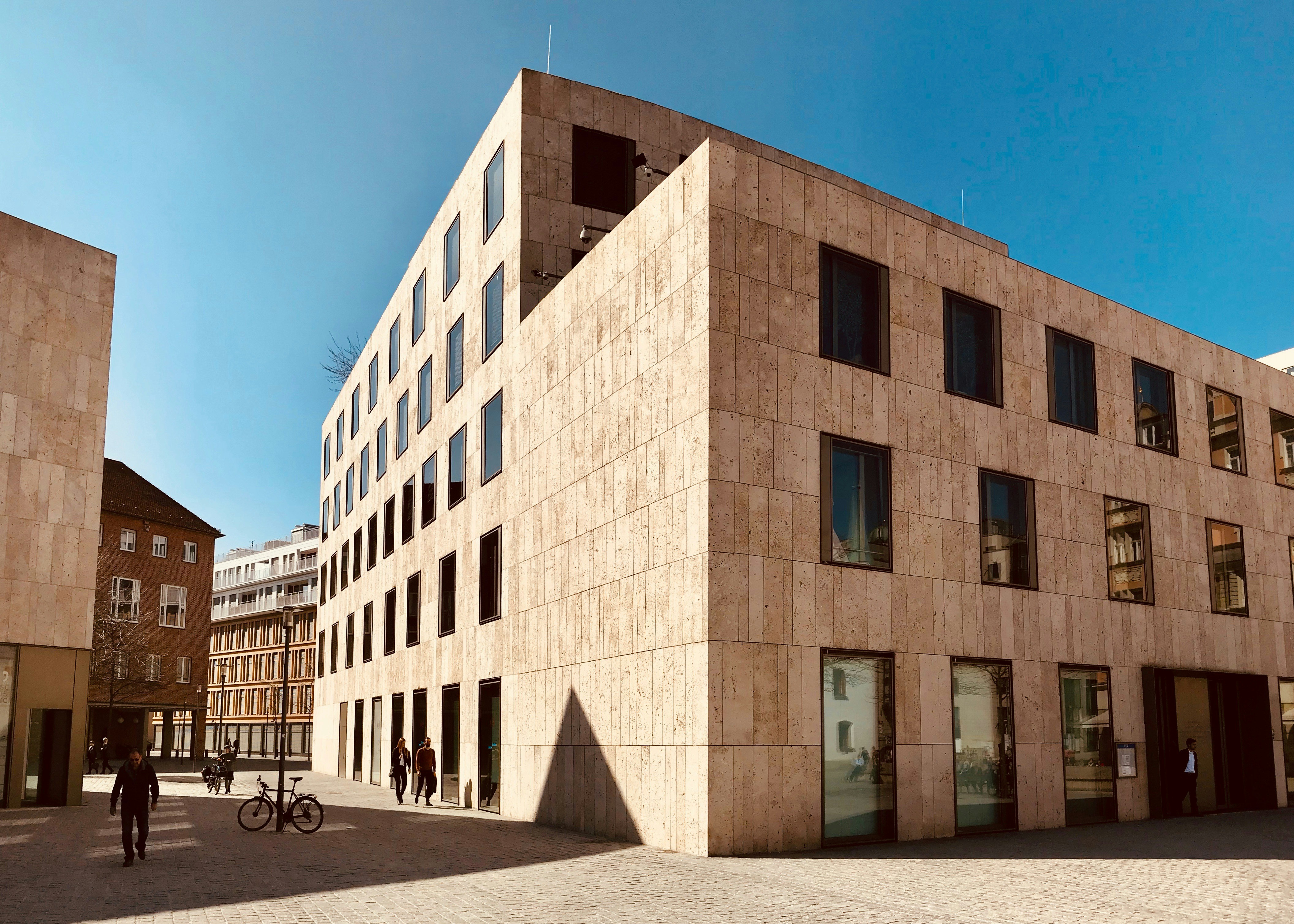 Contemporary building with a textured facade and large windows, set against a clear blue sky. Pedestrians and cyclists navigate the urban space.