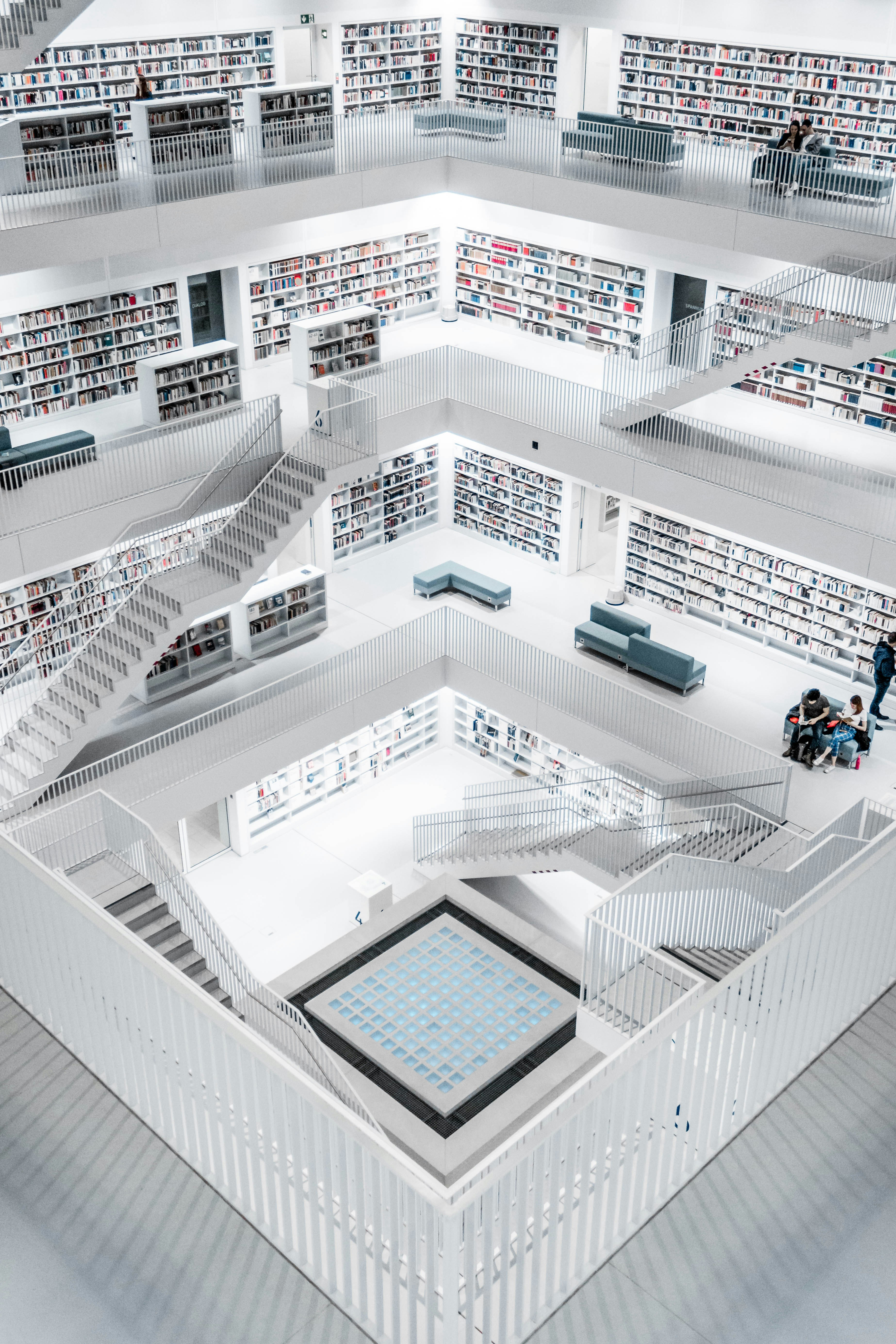Interior view of a modern library showcasing multiple levels of bookshelves, sleek white railings, and a central atrium with a patterned floor. Visitors engage in study and conversation.