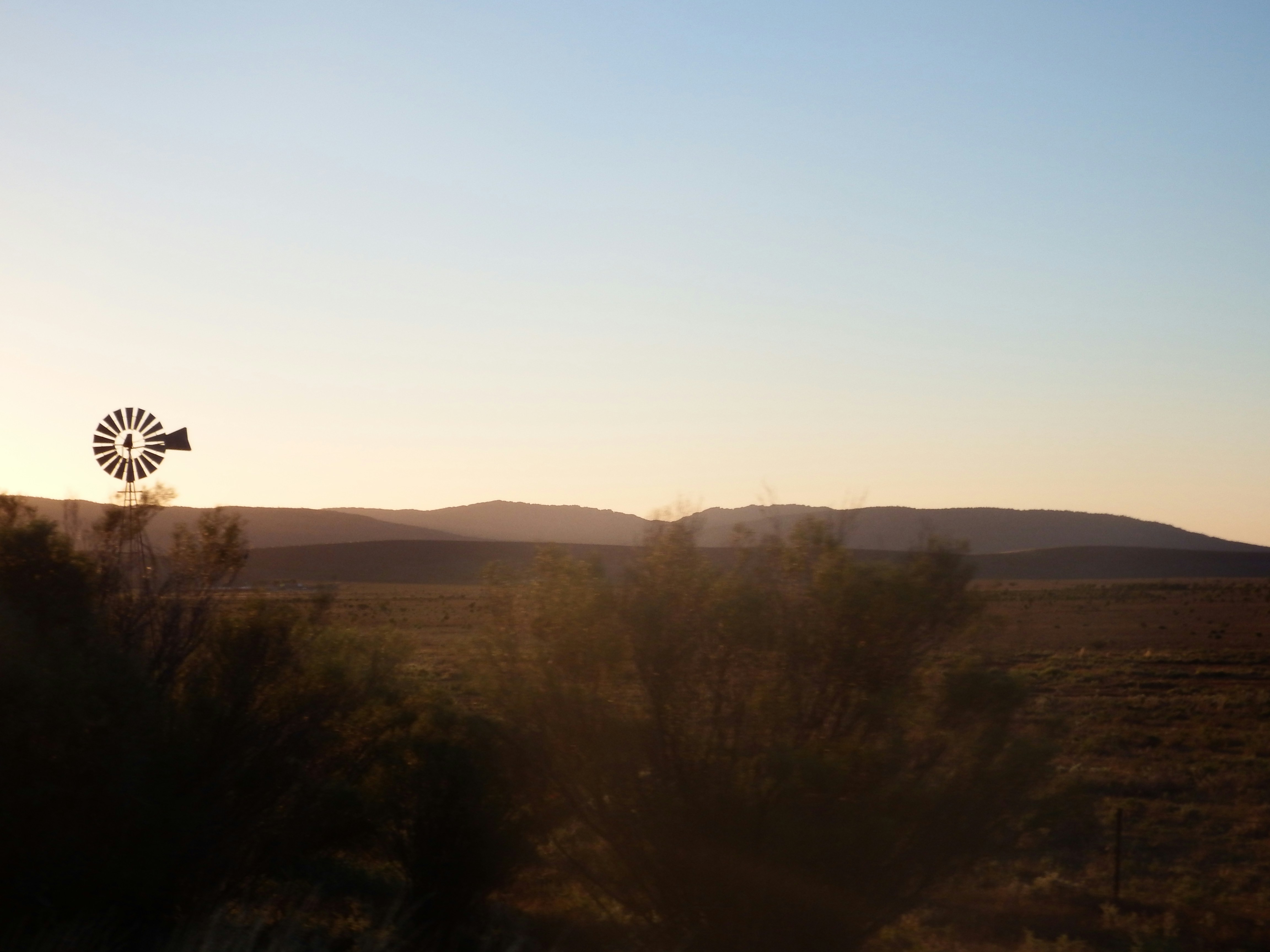 Silhouetted windmill stands against a fading sunset, framed by distant hills and sparse vegetation.