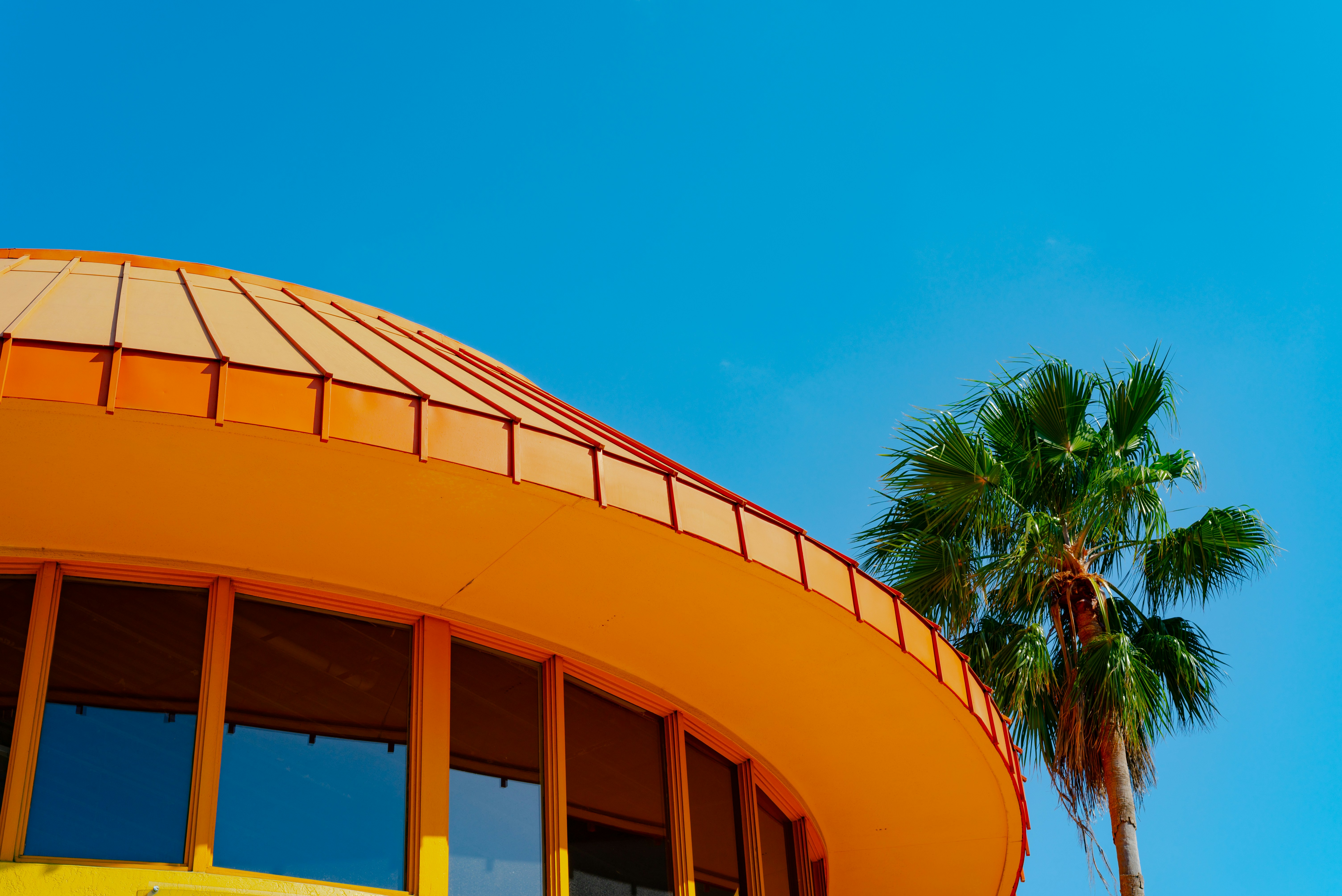 Orange-domed building with glass windows set against a vivid blue sky and a palm tree.