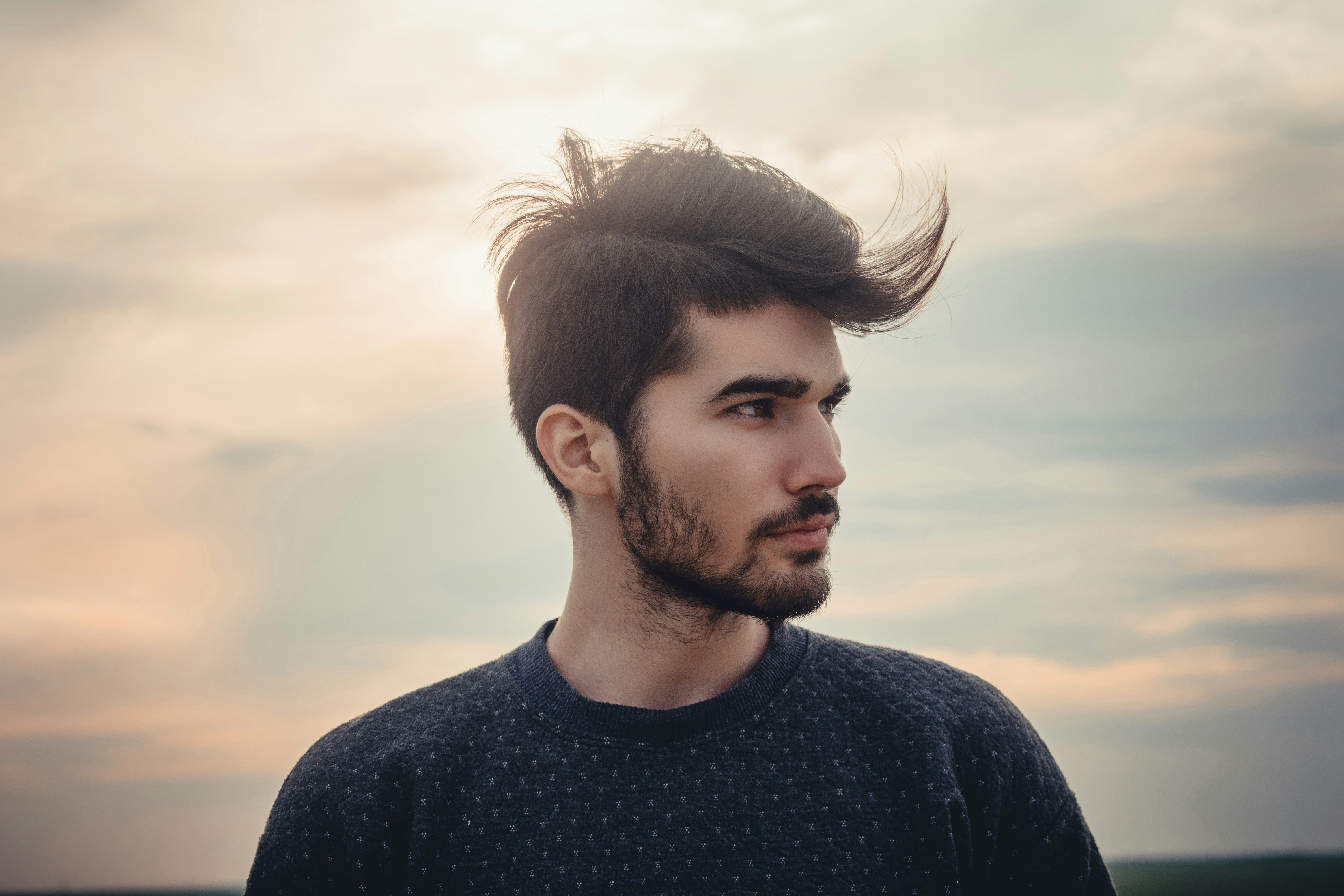 Young man with styled hair gazing thoughtfully against a cloudy sky backdrop.