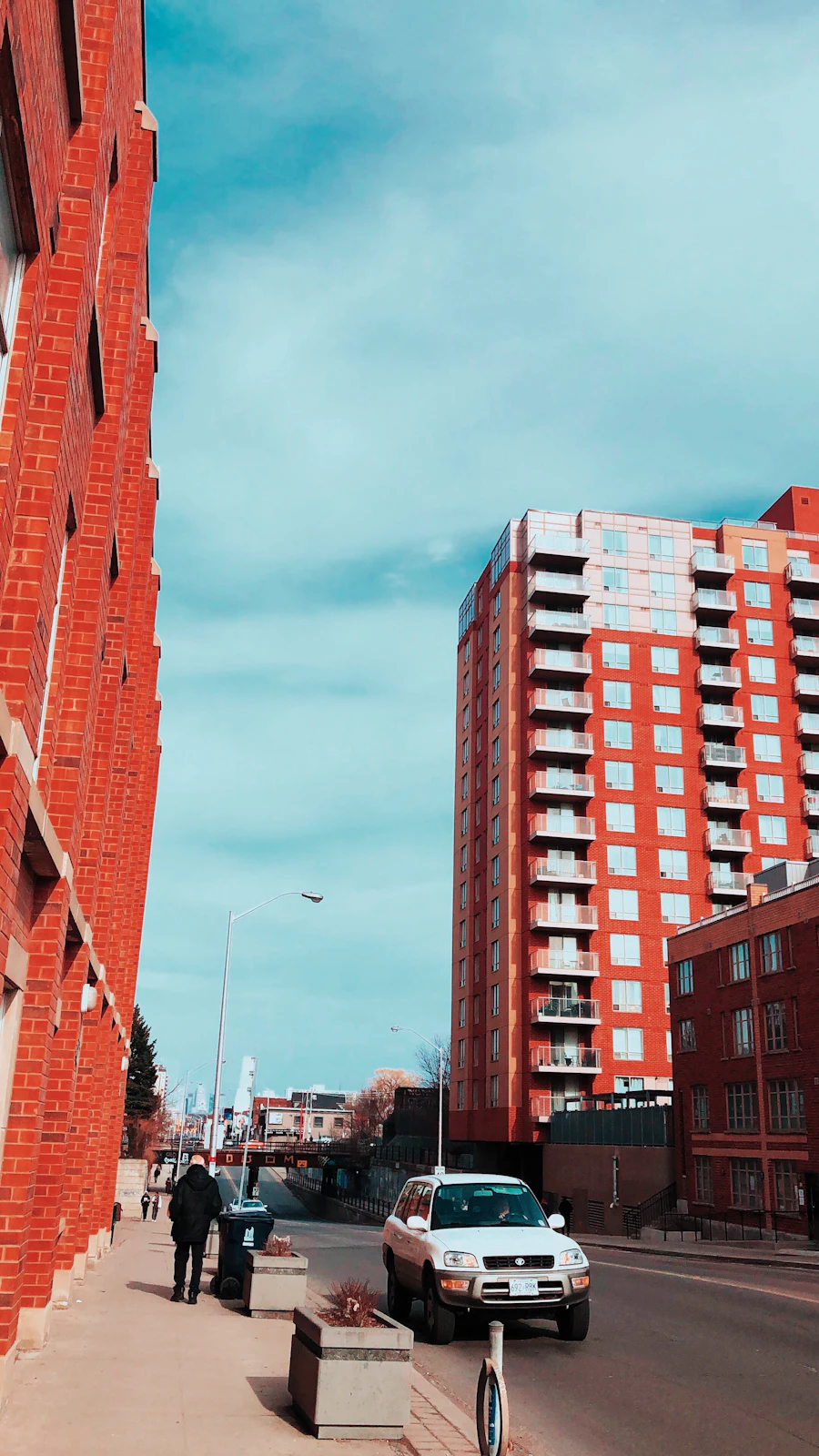 Urban street lined with red brick buildings, a white SUV parked along the curb, and a lone pedestrian walking on the sidewalk beneath a bright cyan sky.