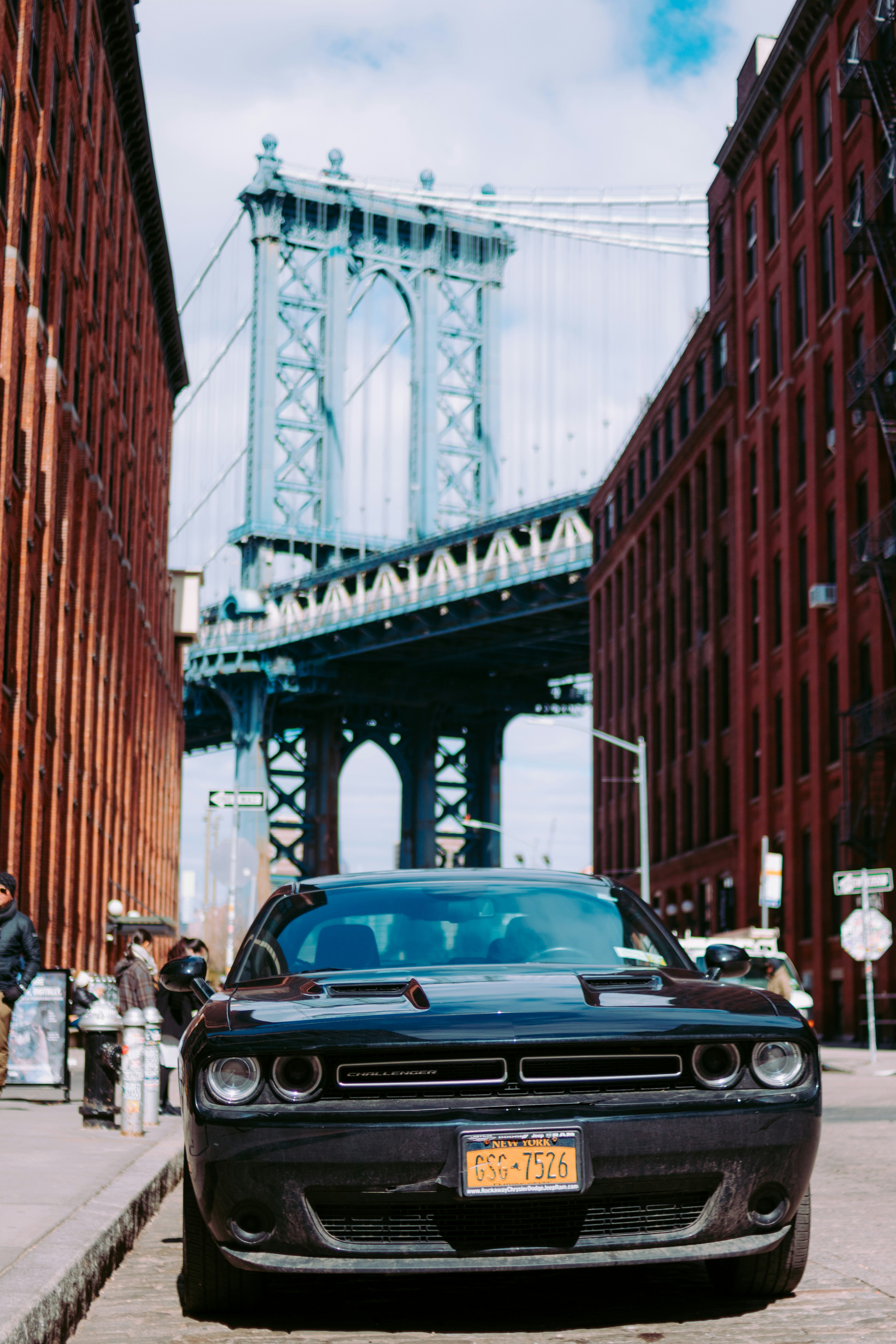 Classic muscle car parked on cobblestone street with the Manhattan Bridge in the background, framed by historic brick buildings.