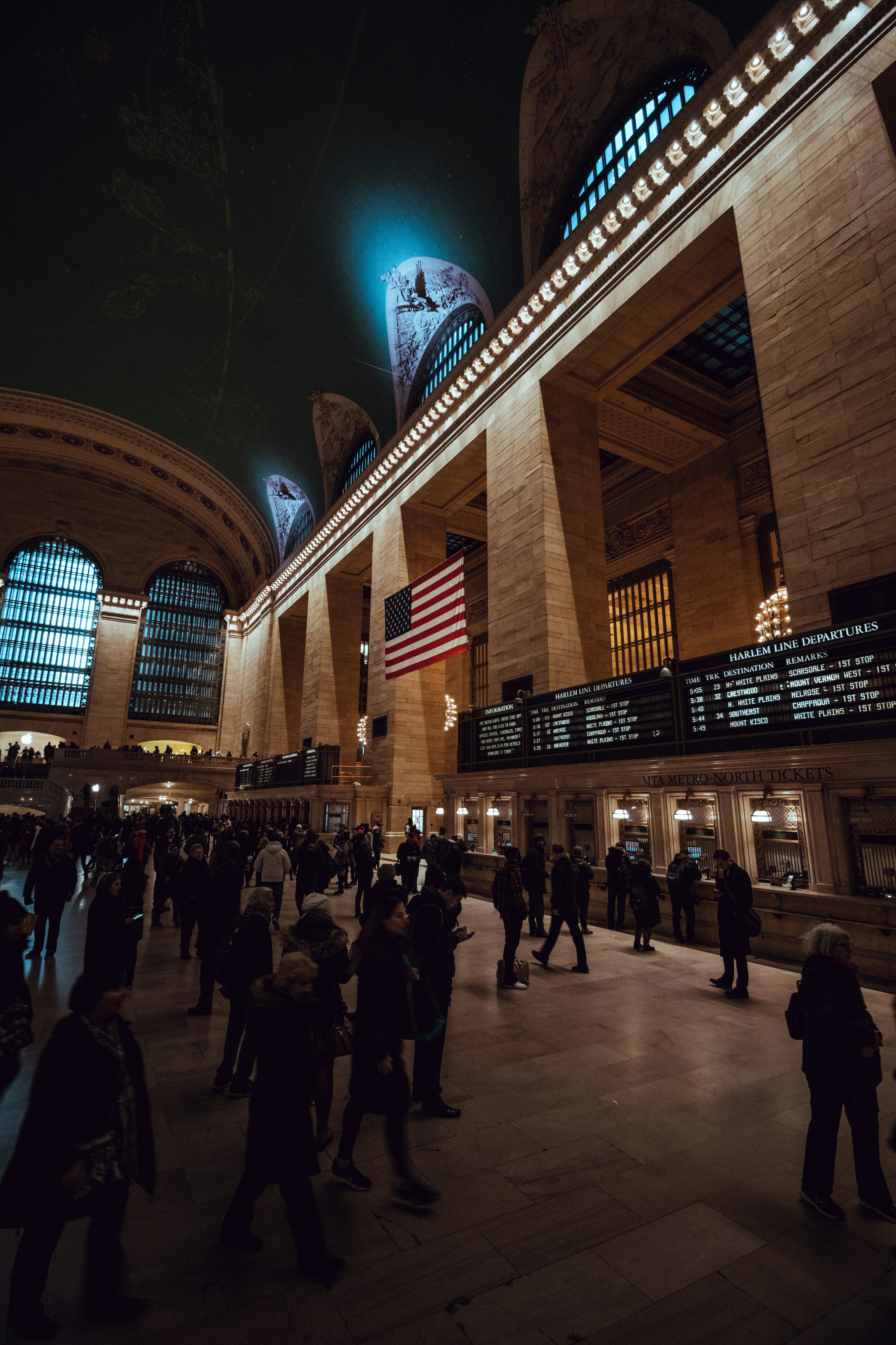 Grand Central Terminal's iconic architecture adorned with an American flag and illuminated departure boards, bustling with travelers. The high ceilings and large windows create a grand atmosphere.