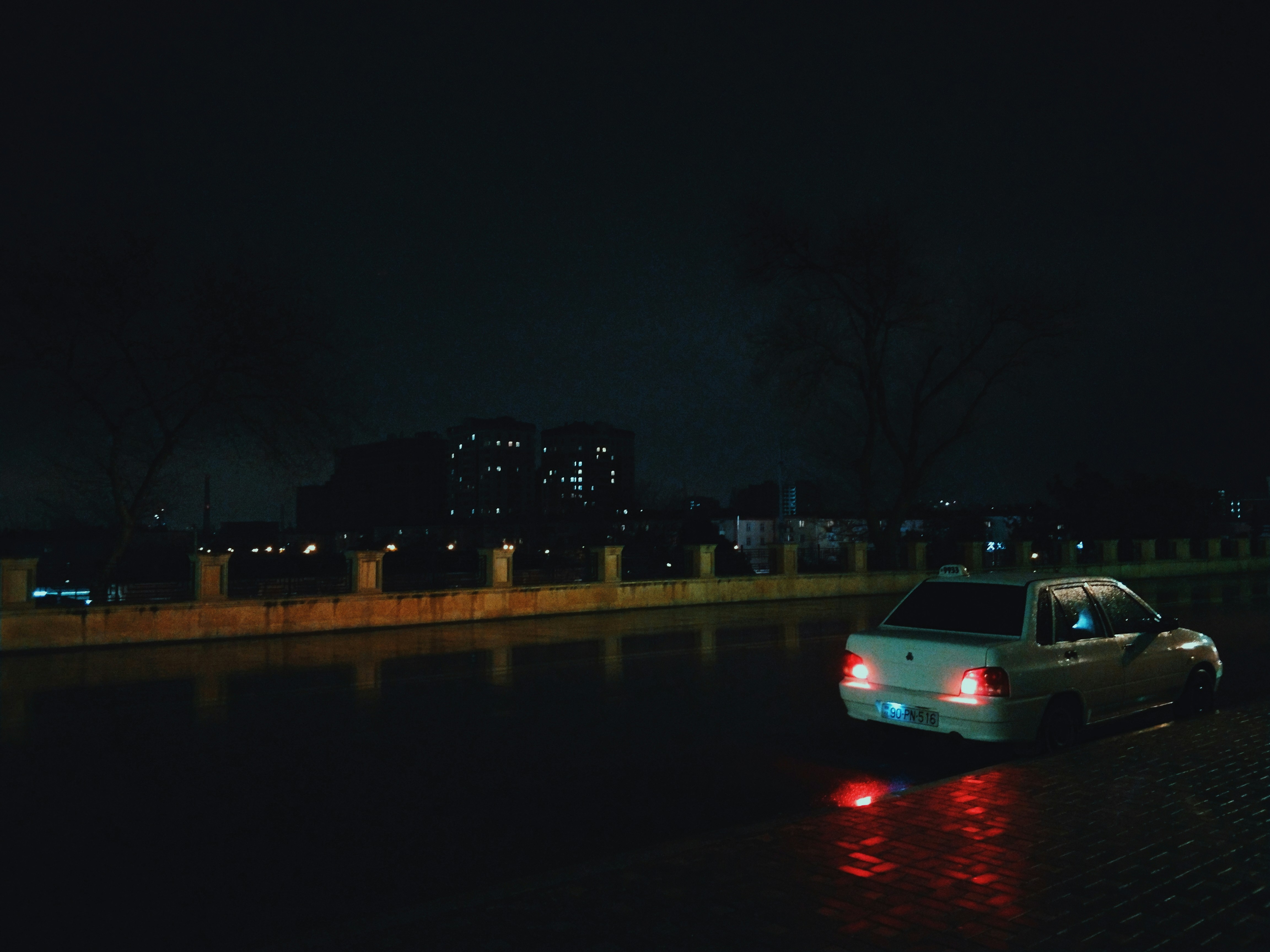 White car with glowing tail lights parked by a dark, rainy riverside with city lights in the distance.