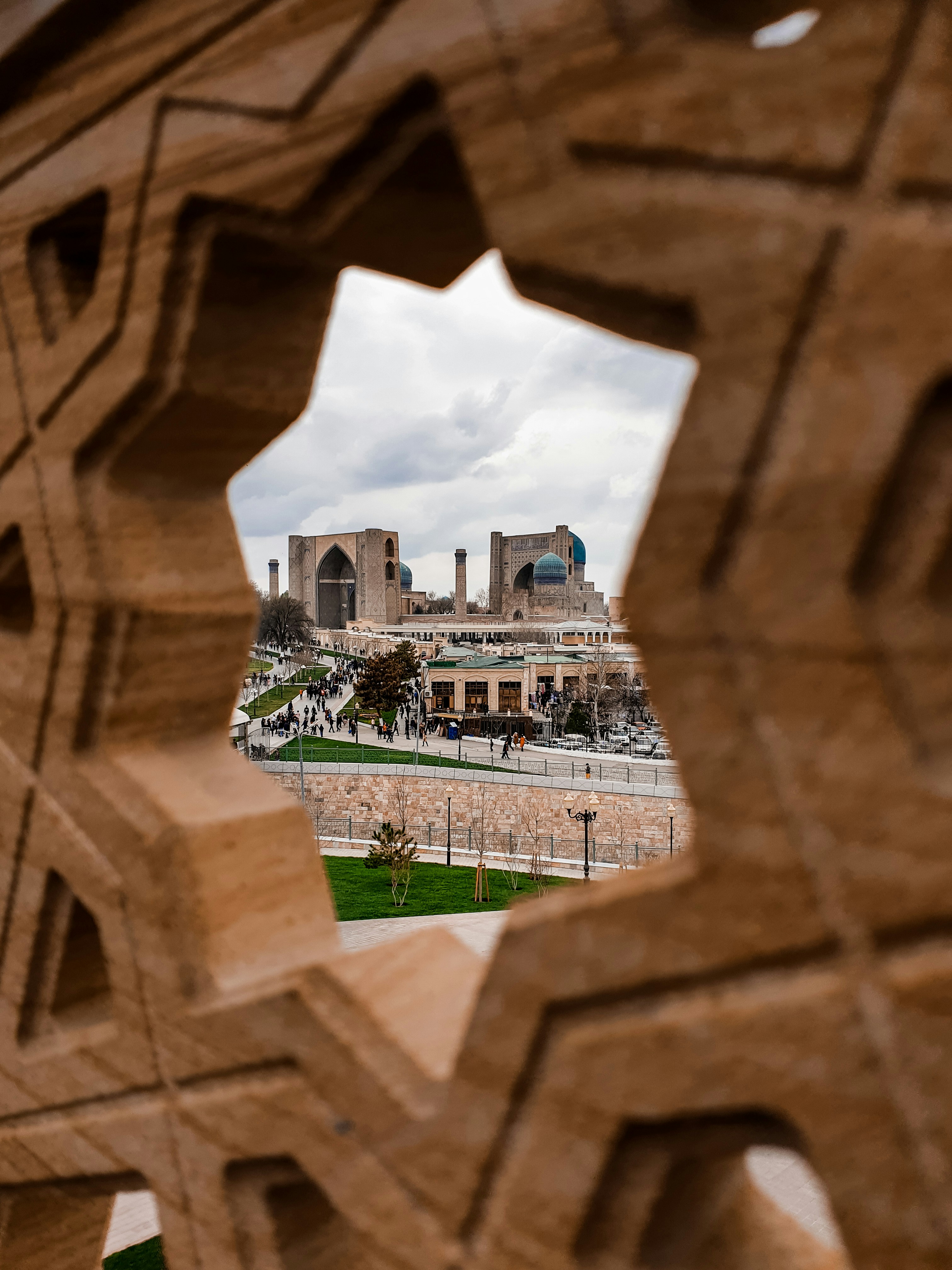 Star-shaped opening in a weathered stone wall frames a distant city plaza with turquoise domes and a green park.