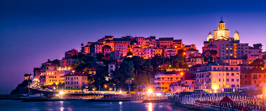 Colorful hillside town glowing at twilight above calm water, with a brightly lit church on the right and reflections along the waterfront. Purple-blue sky fades into warm city lights.