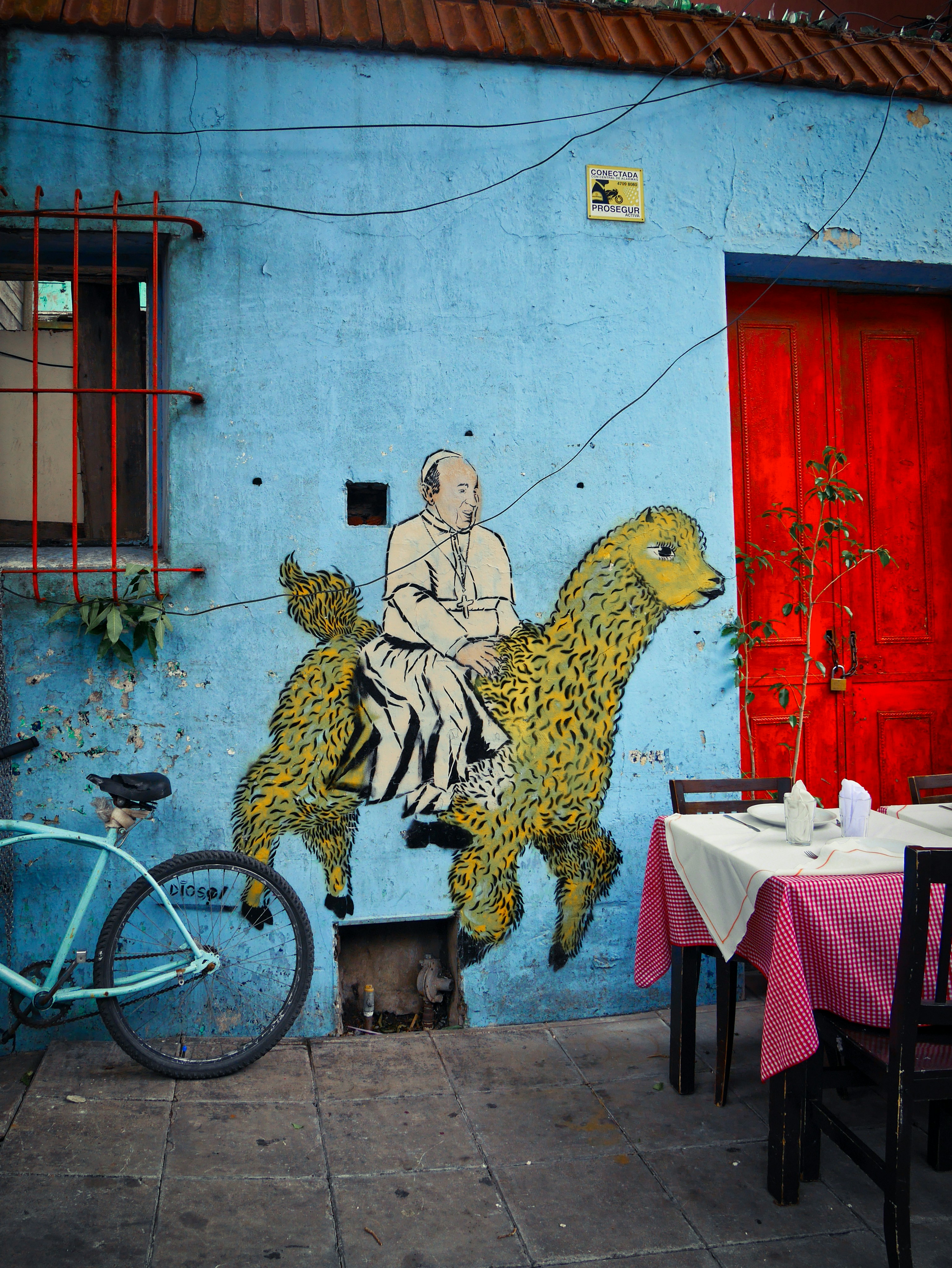 Mural painting on a vivid blue wall depicts a man in white robes riding a golden, patterned camel, creating a focal narrative in a bustling street scene. A turquoise bicycle, a red door, and a table with a pink checkered tablecloth add urban context.