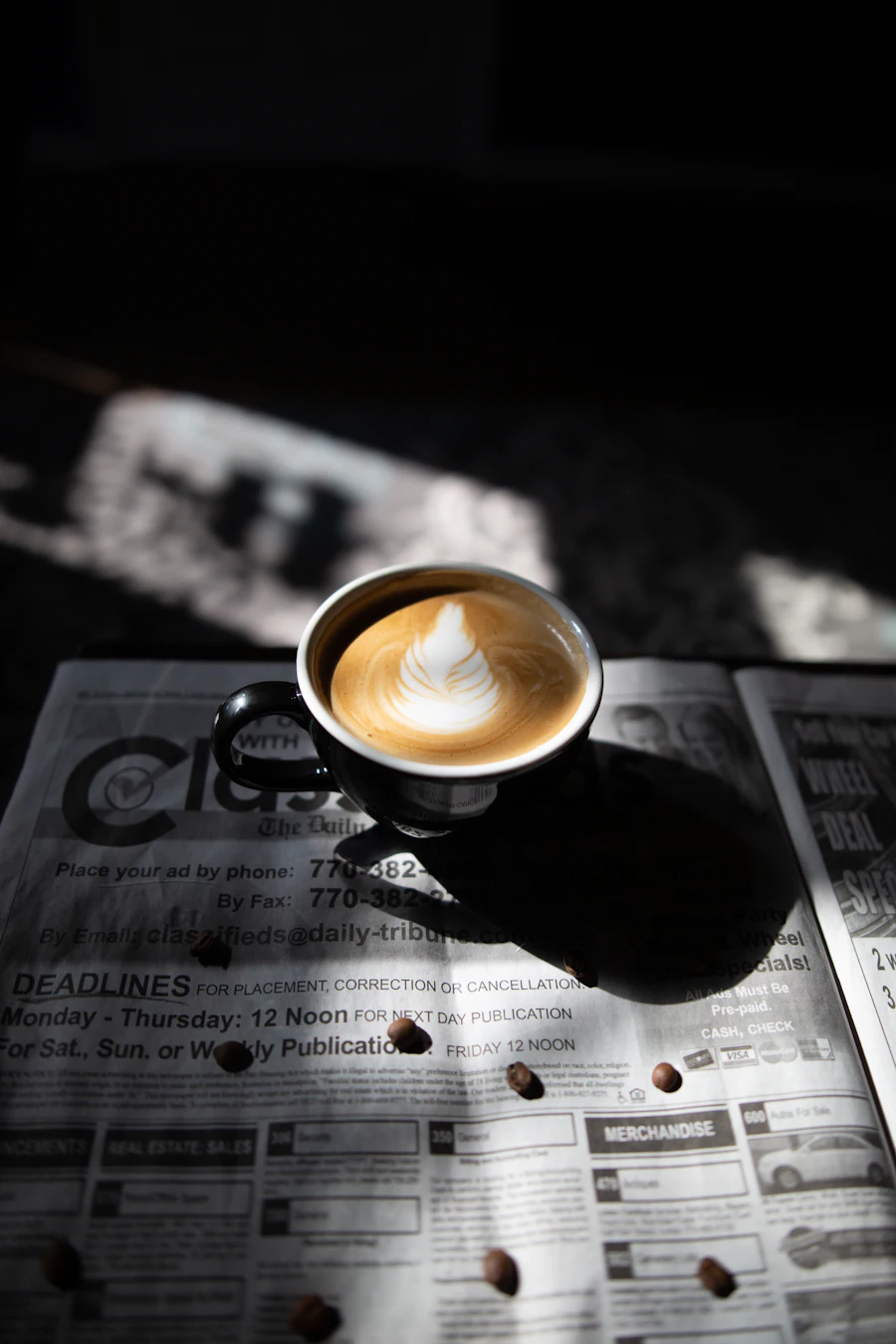 A cappuccino with a leaf-shaped latte art sits on a newspaper spread, lit by a strong patch of sunlight. Coffee beans are scattered across the page, casting a deep shadow.