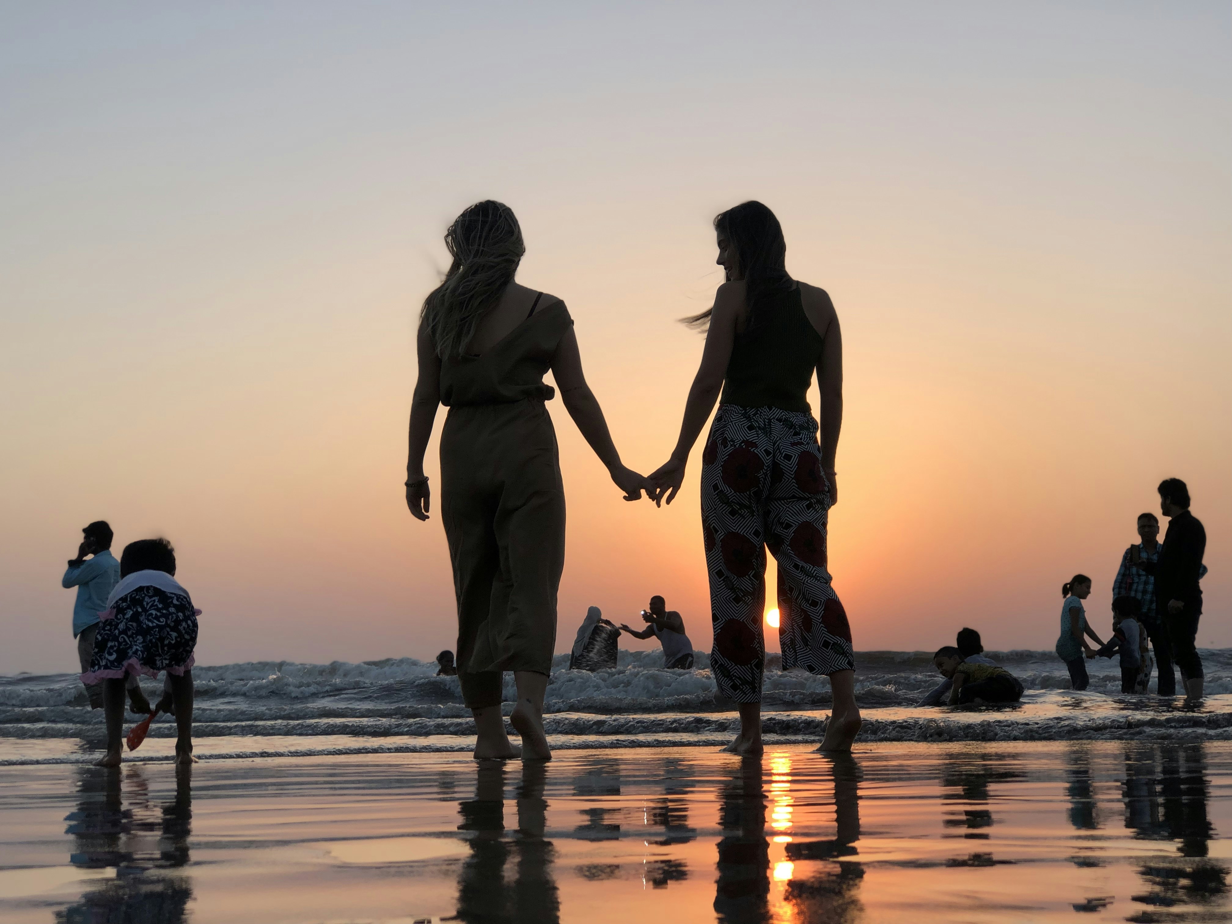 Two people holding hands walk along the beach at sunset with others in the background and reflections on the wet sand.