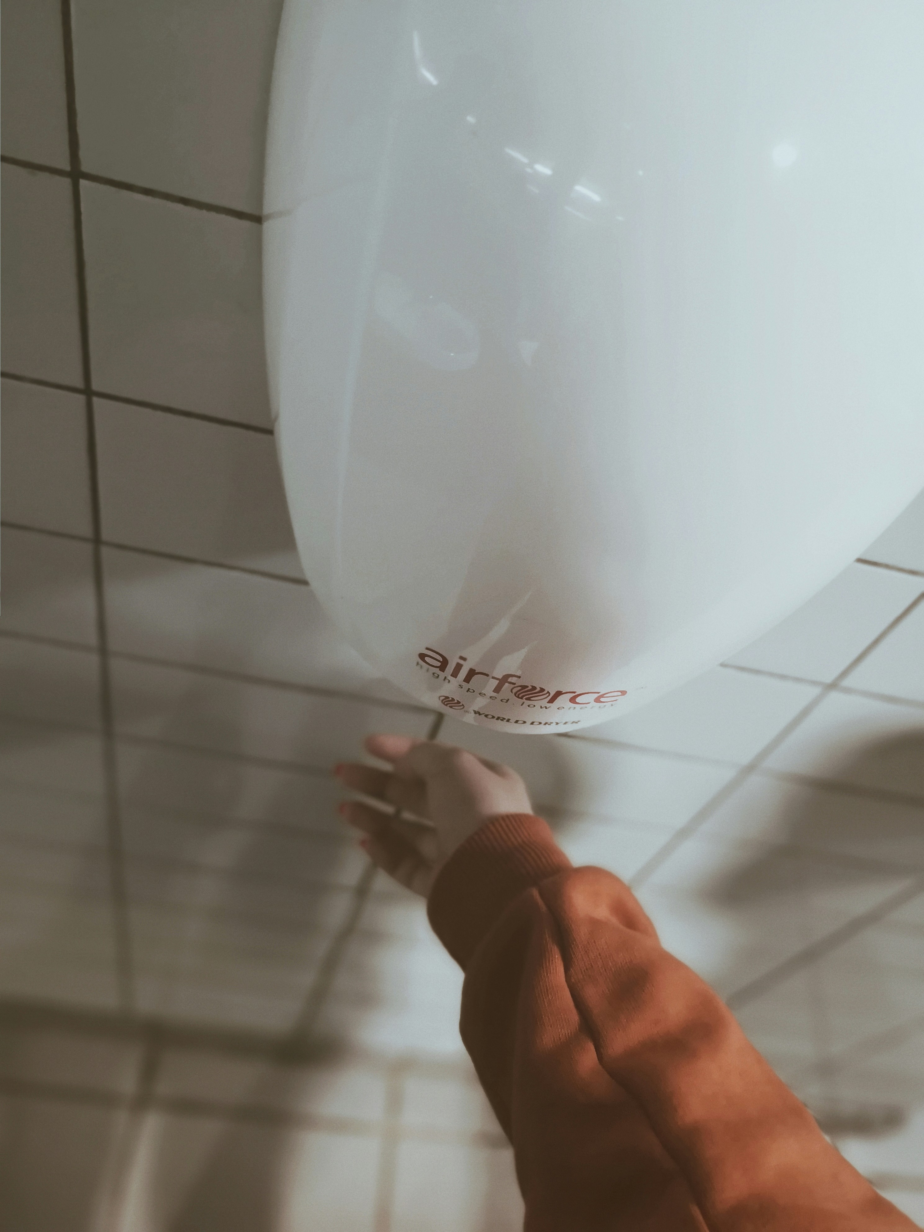 Orange-sleeved reach toward a gleaming air dispenser in a tiled bathroom