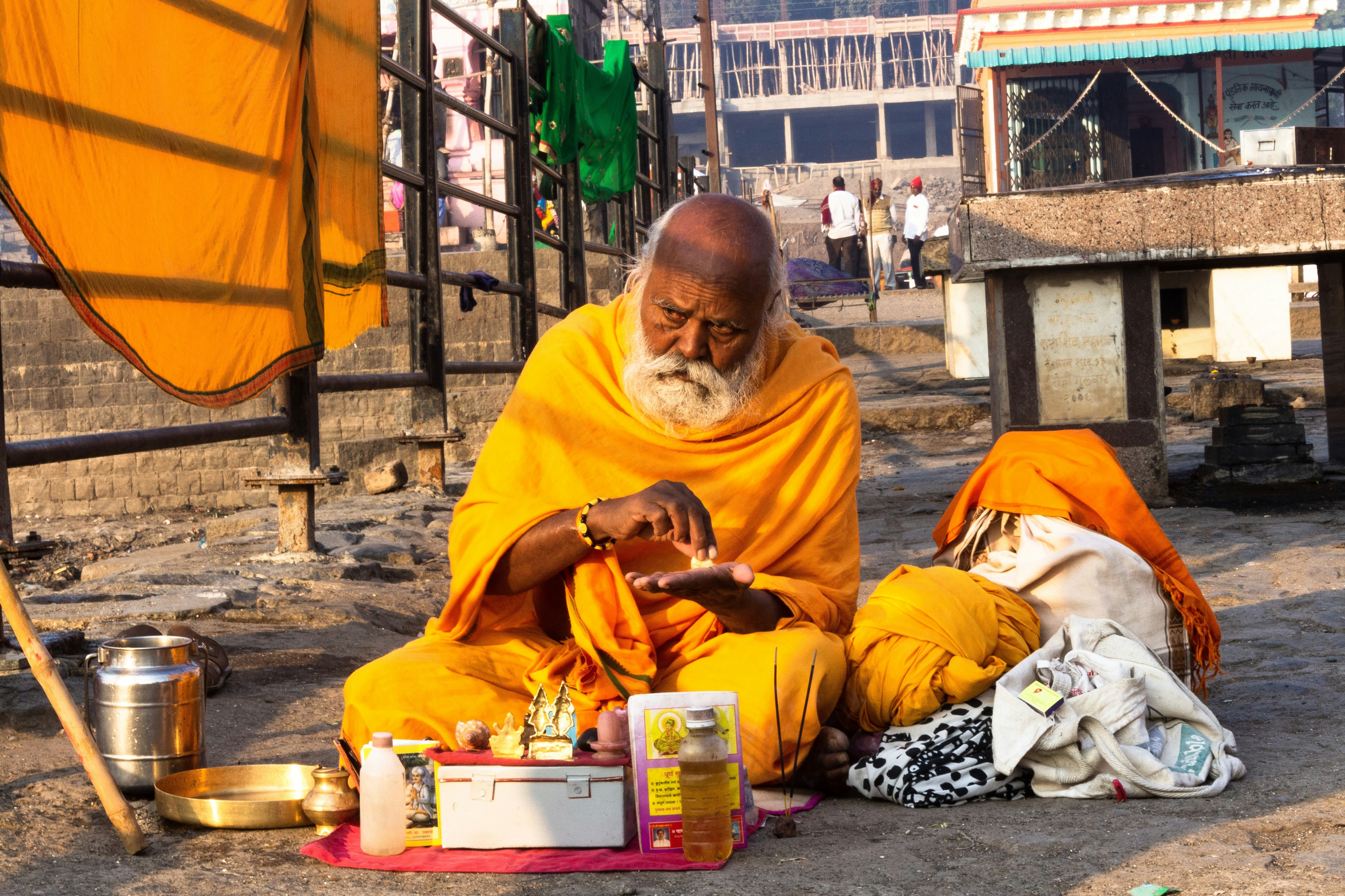 Elderly man in orange robes sits by the riverbank, surrounded by ritual items in the morning light.
