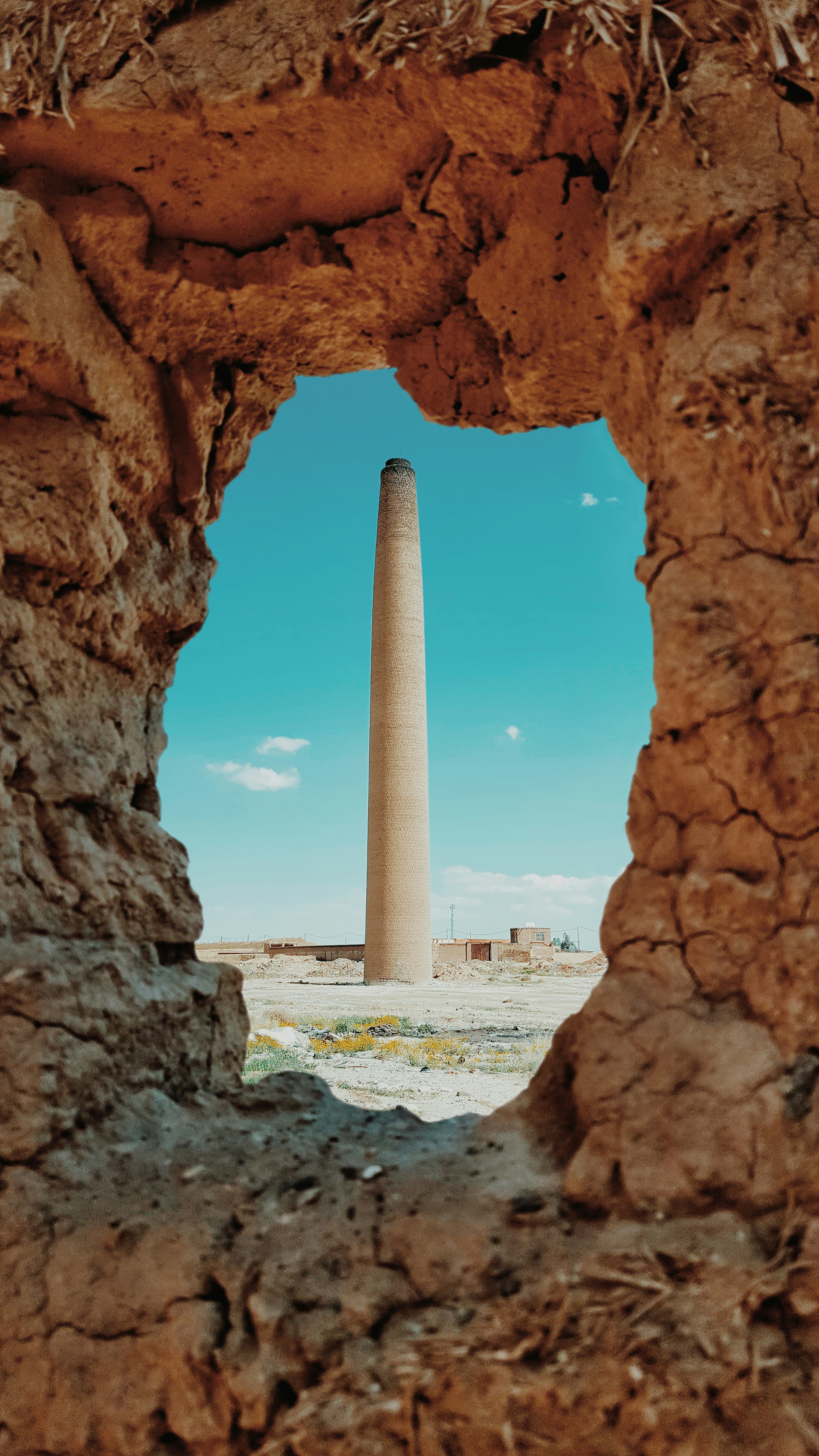 Desert landscape framed by a rugged clay arch. A solitary cylindrical chimney rises on the horizon under a turquoise sky.