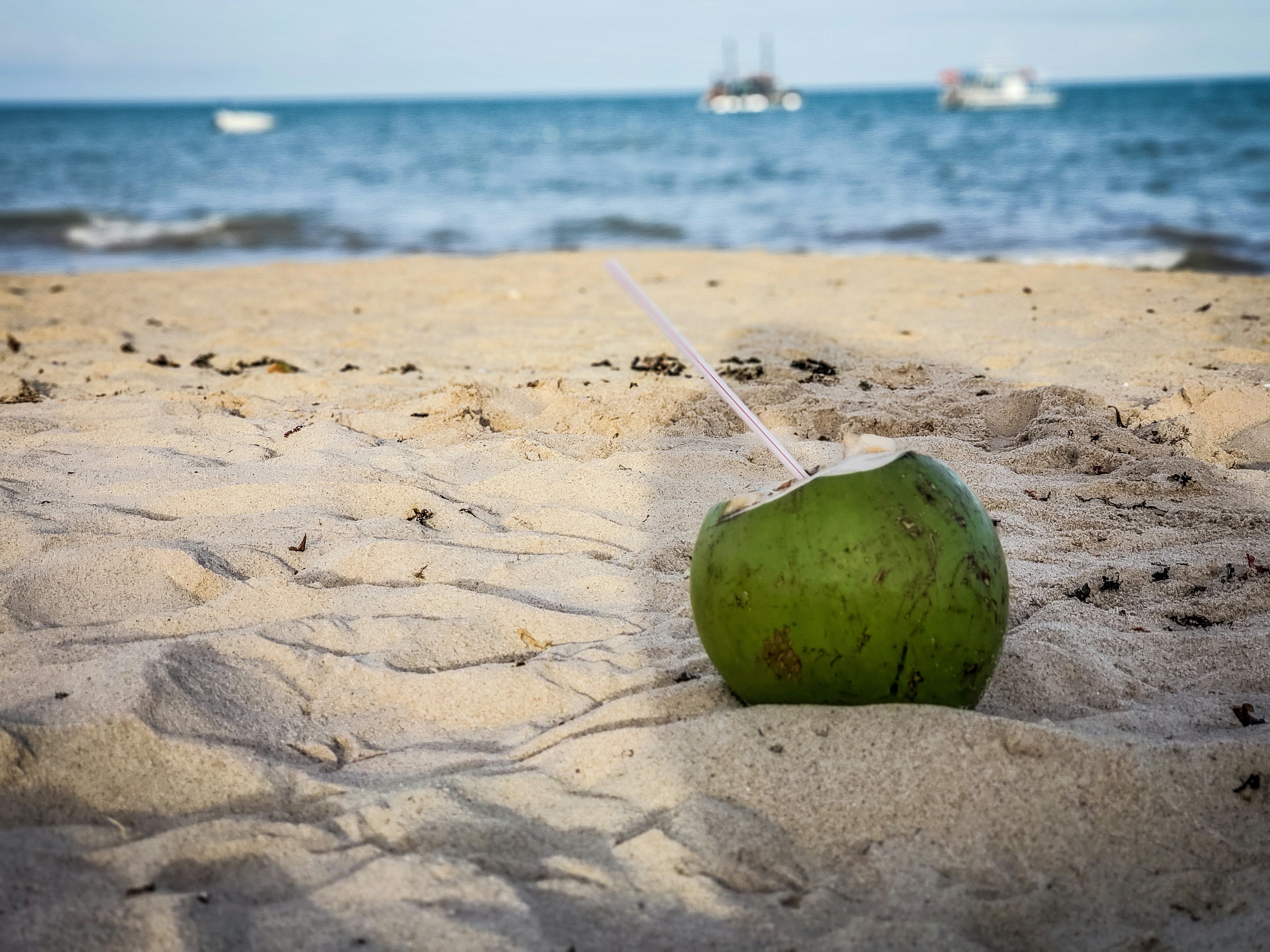 Fresh green coconut with a straw rests on sandy beach with ocean waves in the background.