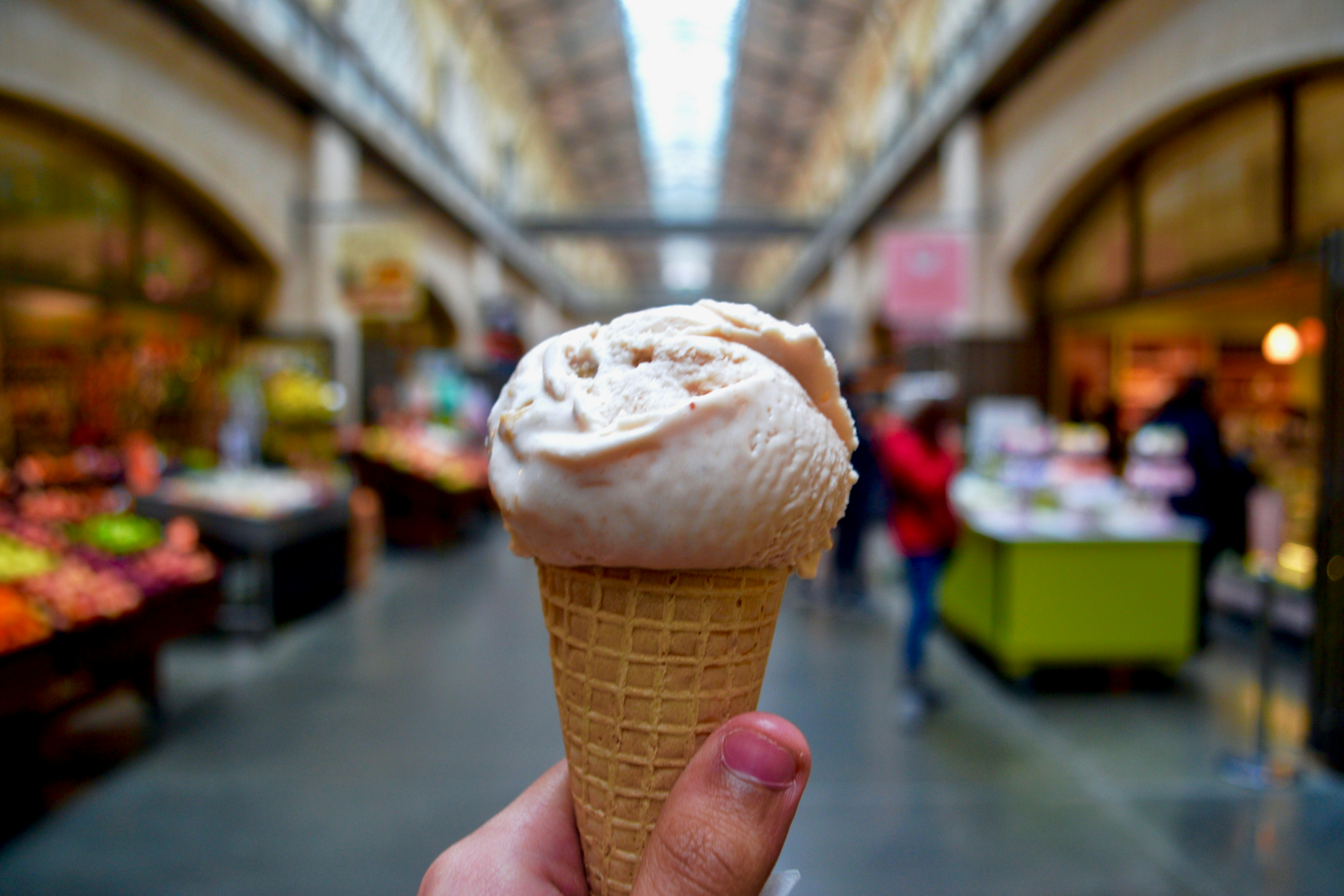 Hand holding a creamy ice cream cone in a bustling market filled with vibrant produce and shoppers in the background.