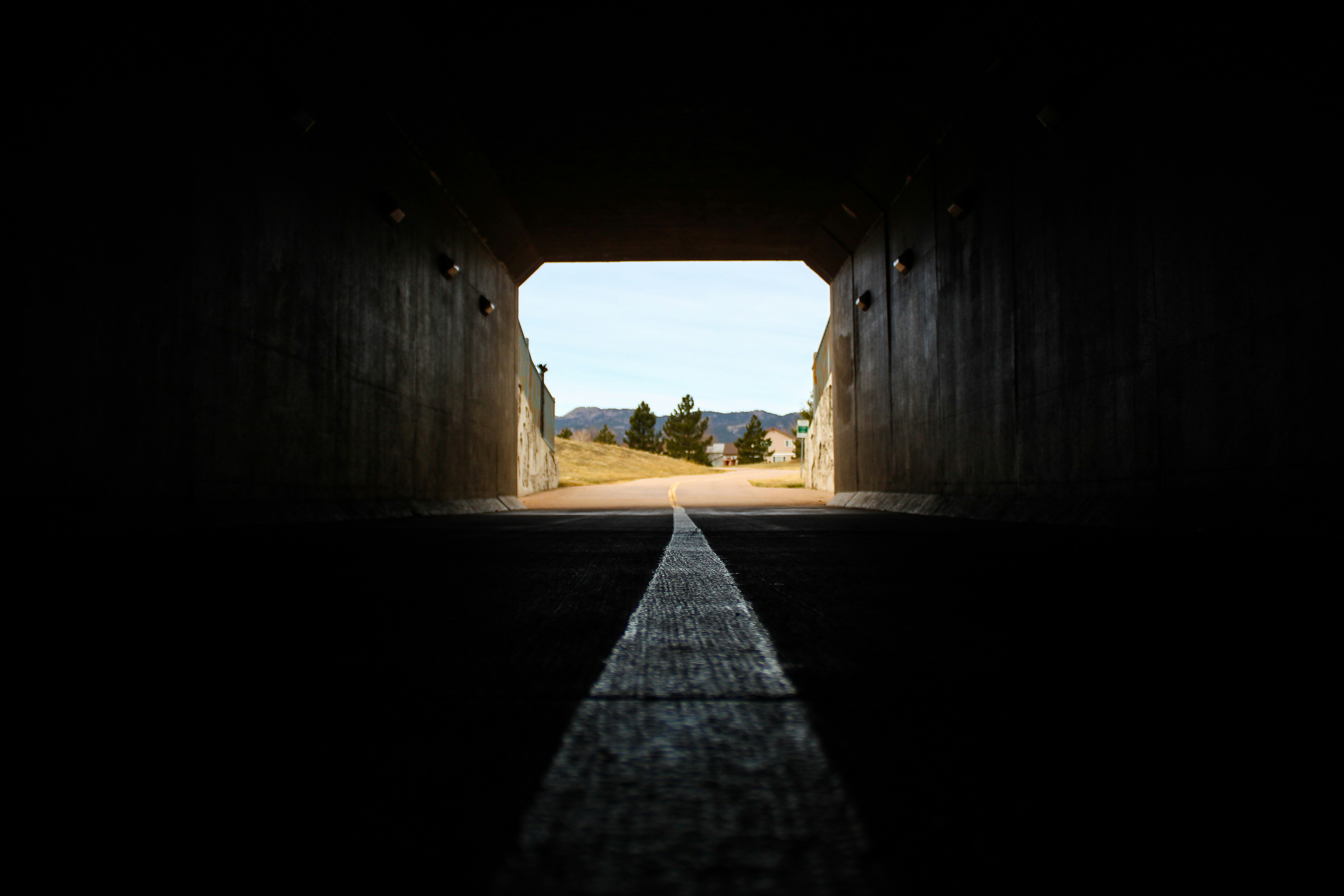 View through a dark tunnel leading to a sunlit bike path with distant trees and blue sky.