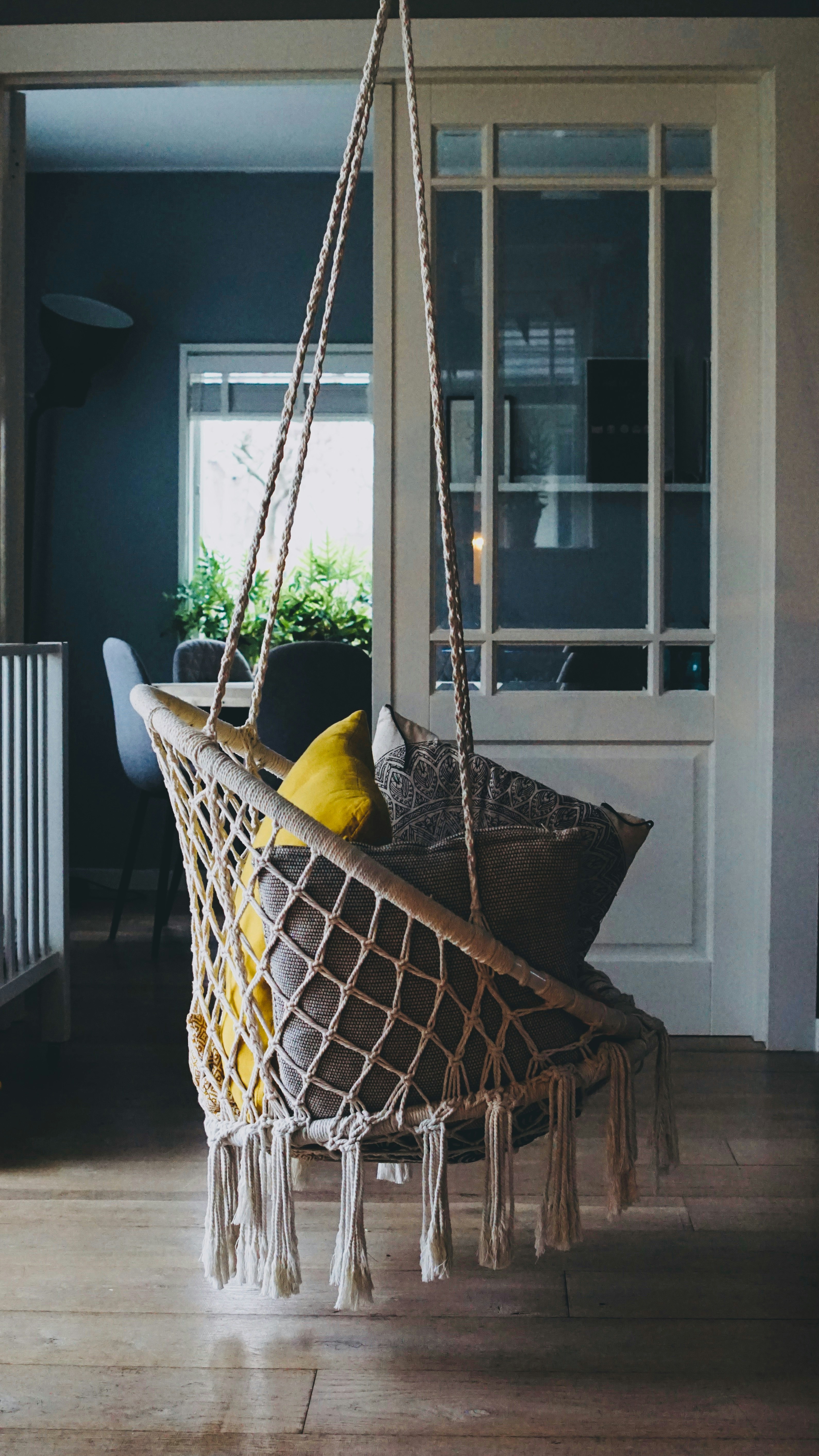 Macrame hammock chair with yellow and gray cushions hangs in a sunlit living room with blue walls and glass-paned doors.