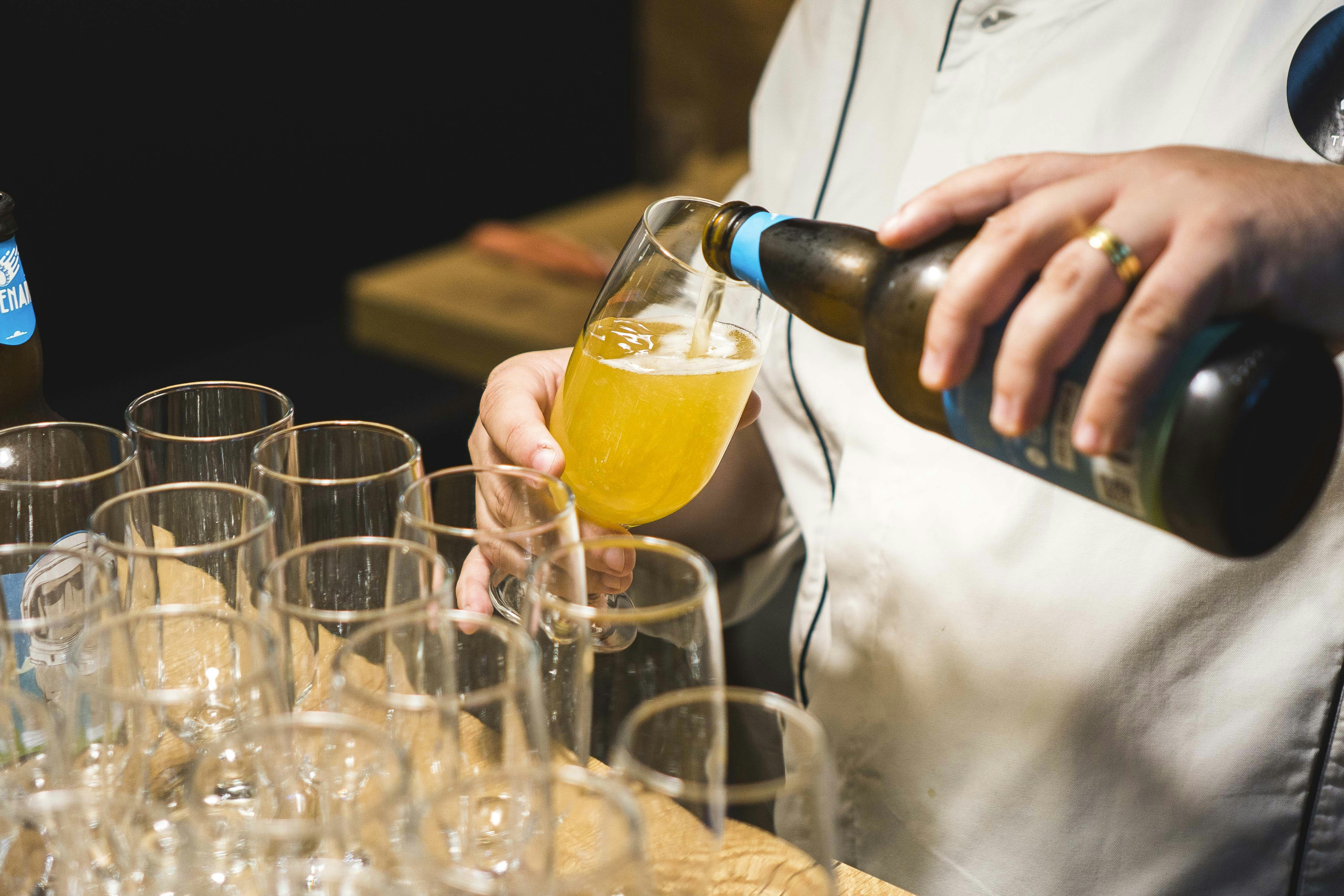 Person pouring beer into a glass surrounded by empty glasses on a wooden counter.