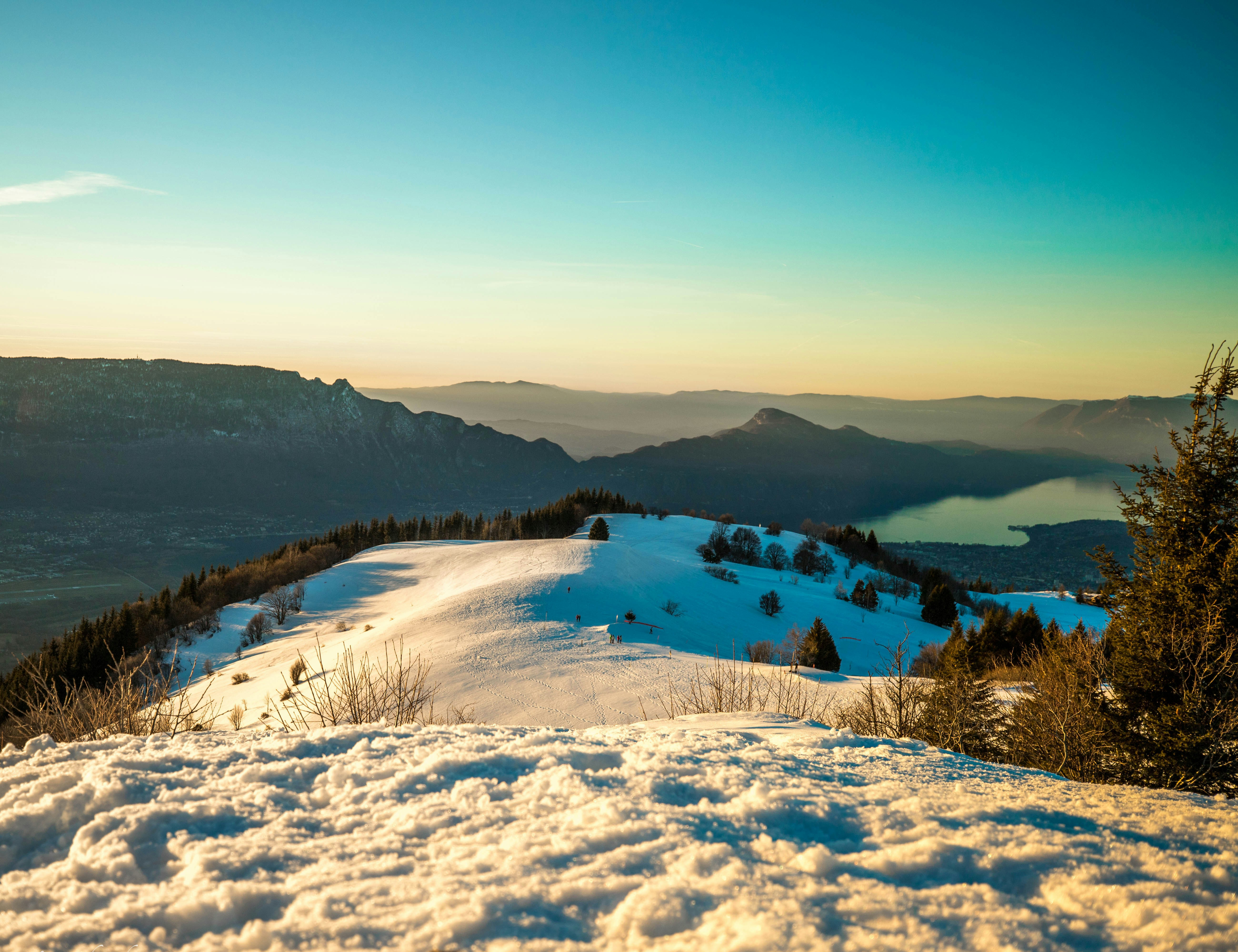 Snow-covered mountain summit overlooking a serene lake and distant hills under a clear blue sky.