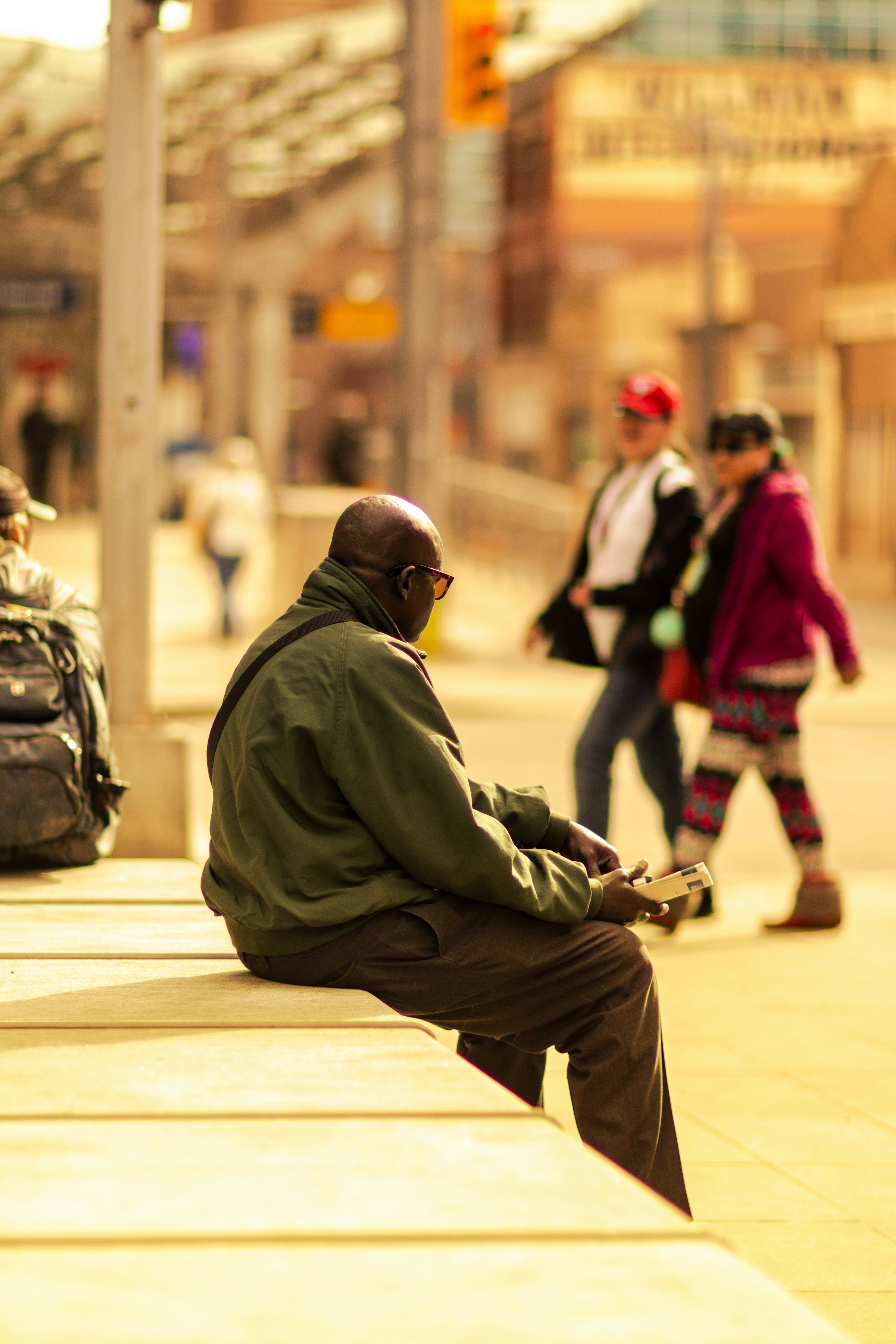 A man seated on a bench, engaged with his phone, amid a lively urban backdrop with passersby. The scene captures a blend of stillness and activity.