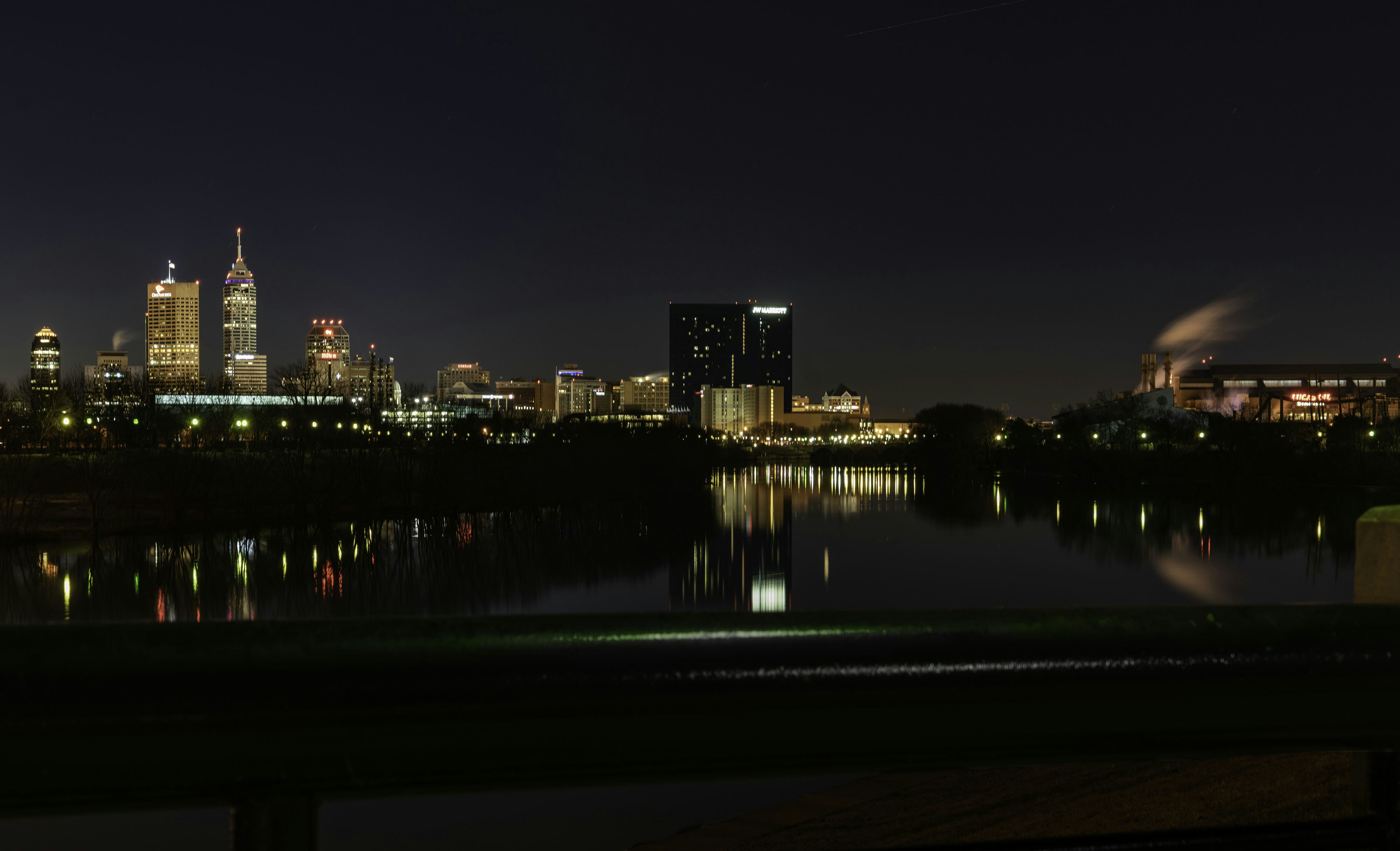 Nighttime city skyline reflected on a calm river with illuminated buildings and dark sky.