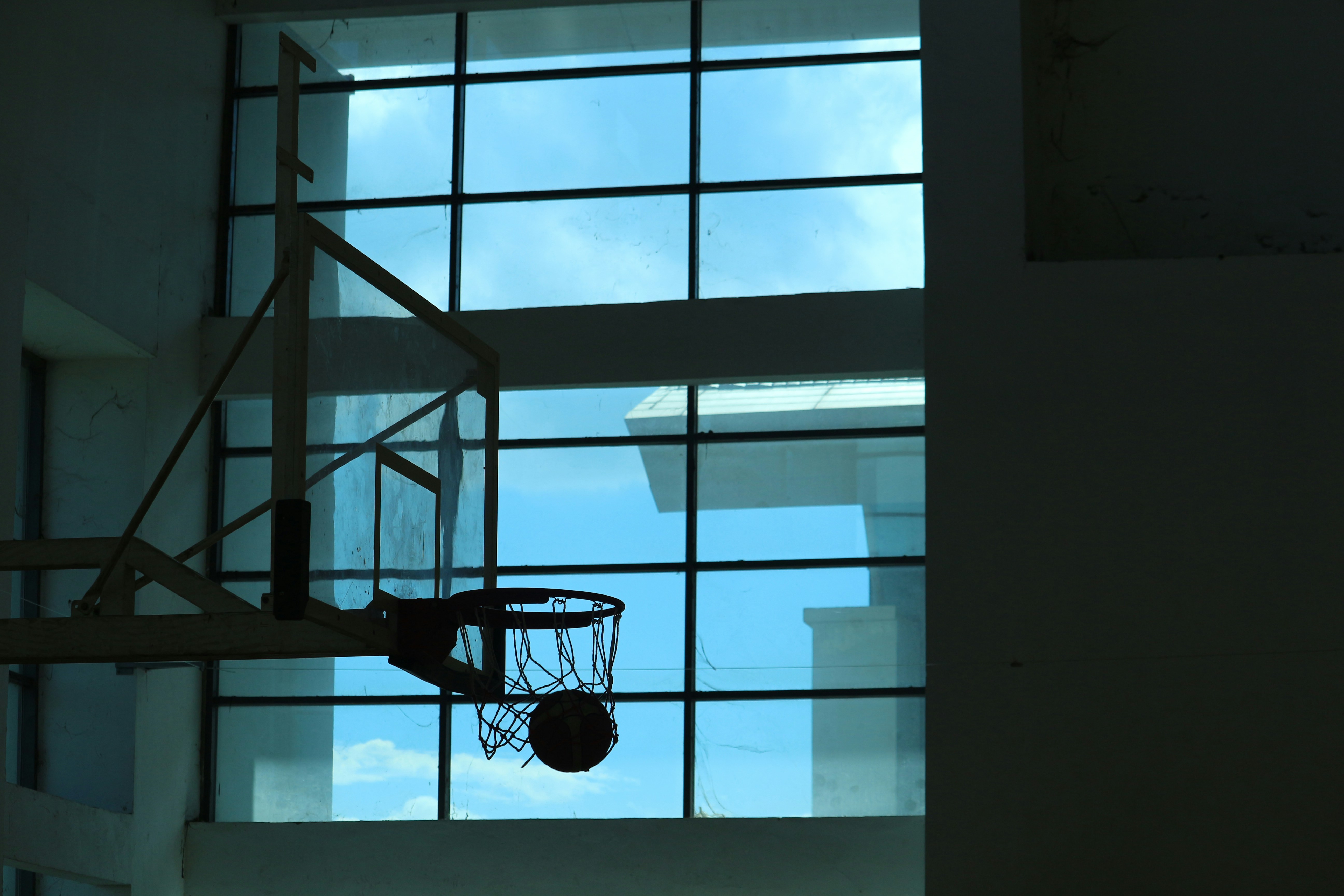 Photograph of an indoor basketball hoop silhouetted against bright blue window panes, with a ball near the net in a dim gym.