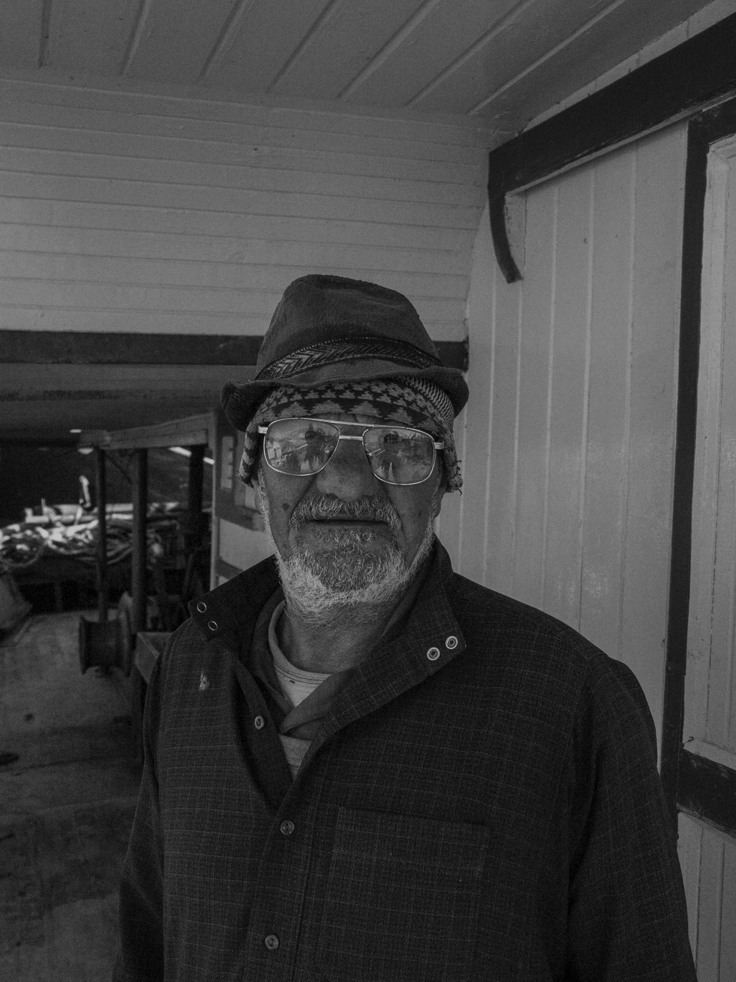 Elderly man wearing a hat and glasses, standing in a rustic interior space filled with hints of history.