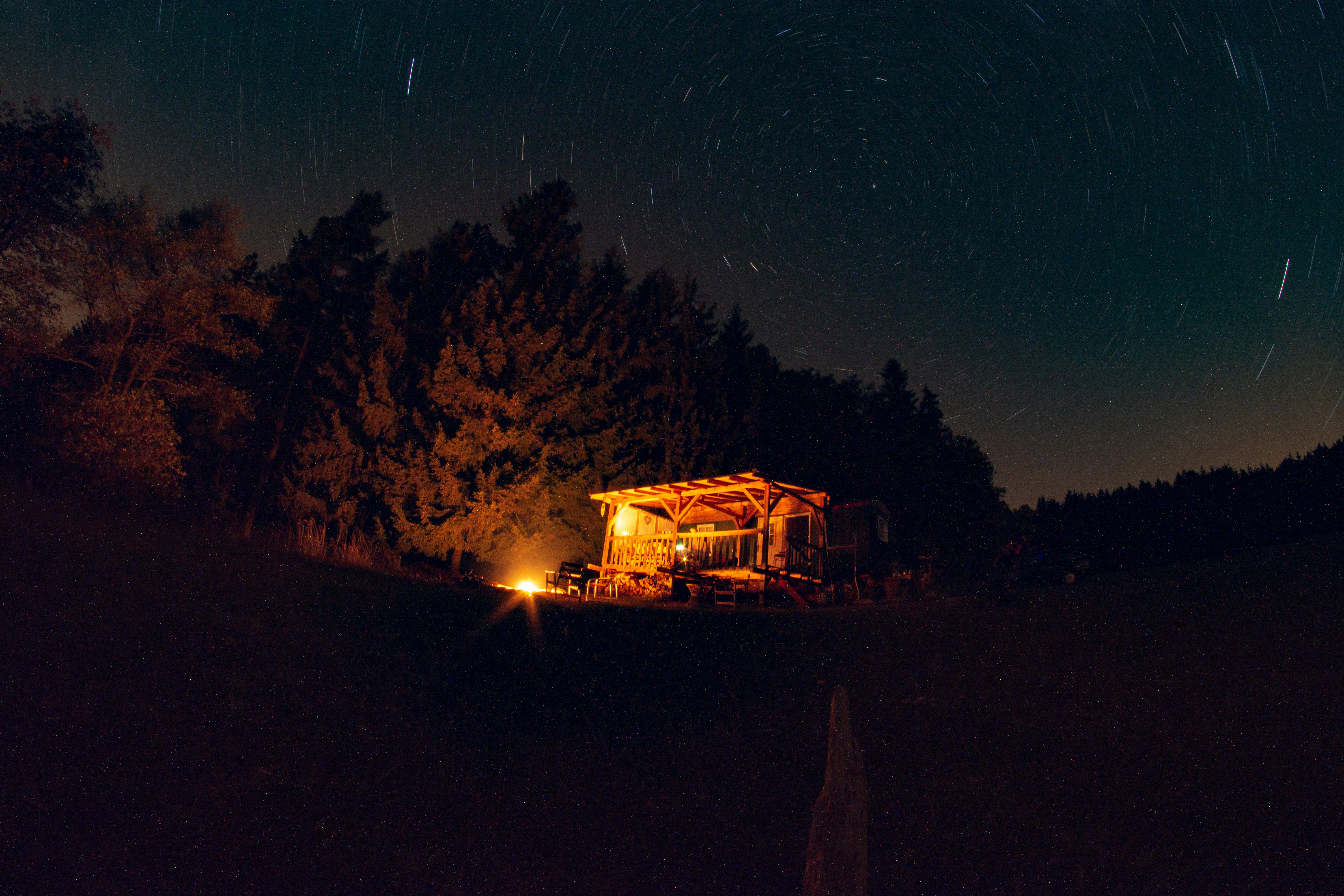 A cozy wooden cabin illuminated by warm light amidst a dark forest, with star trails swirling above in the night sky.