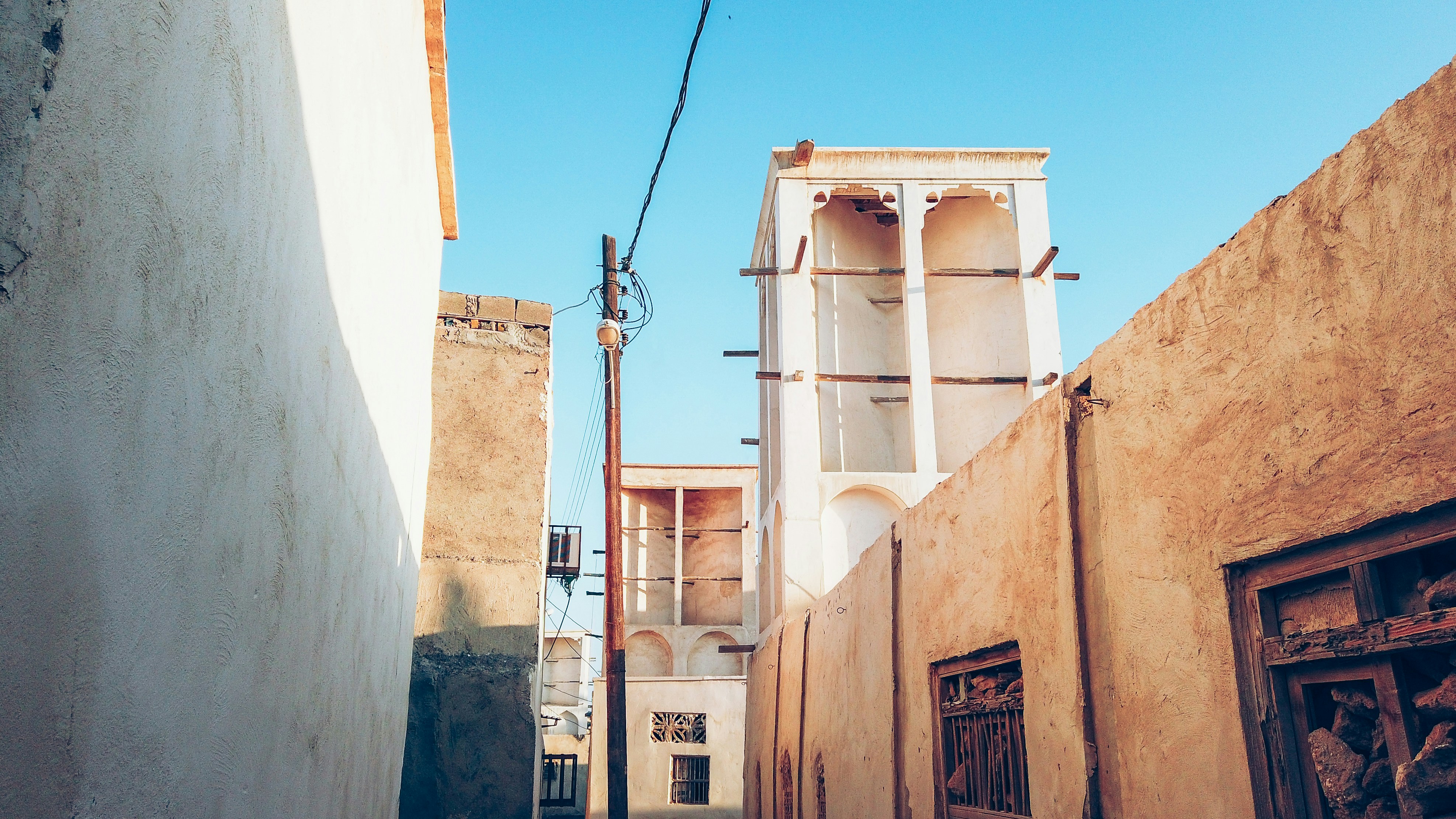 Narrow alley framed by aged walls leads to a traditional windcatcher under a clear blue sky.