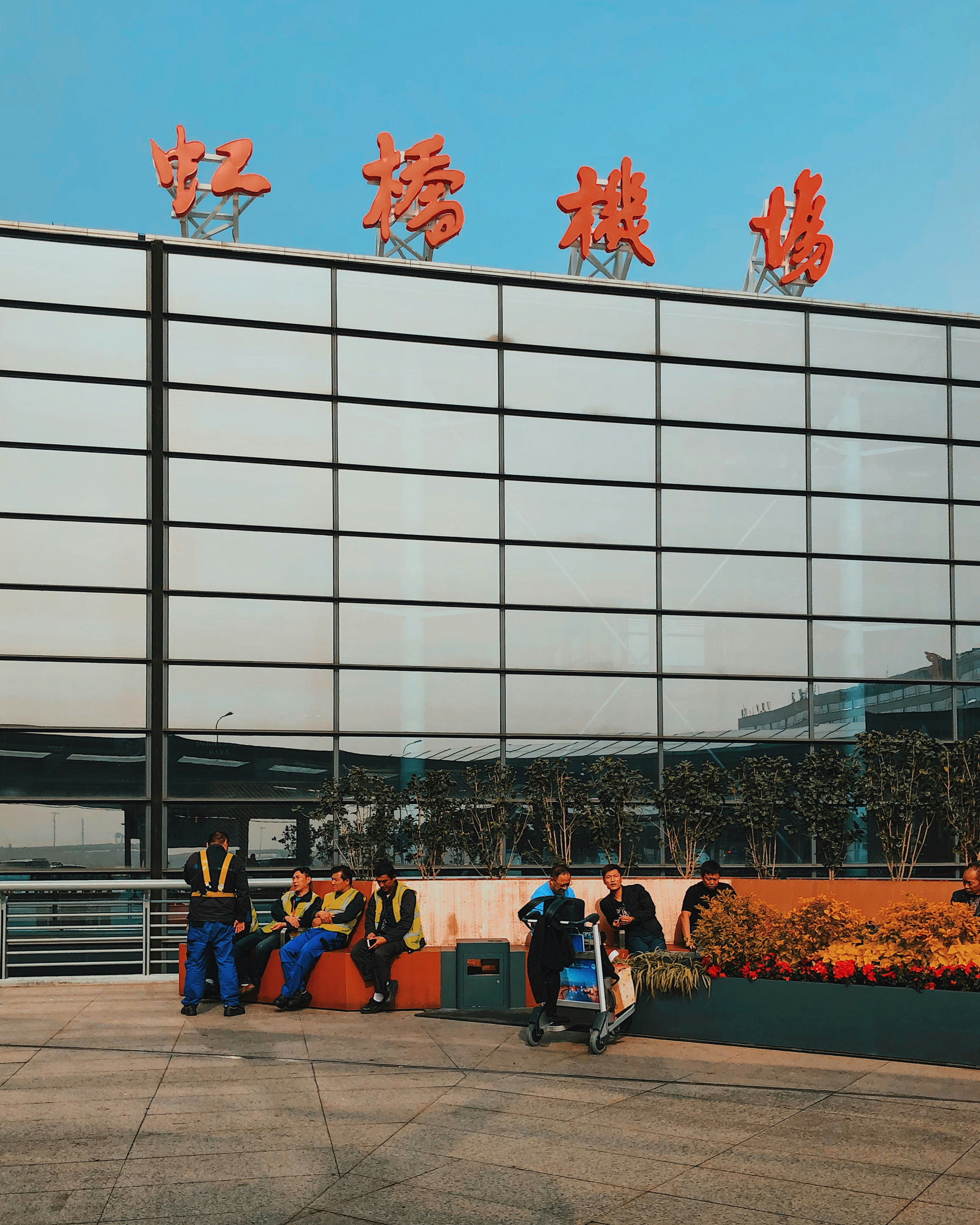 Workers in bright uniforms relax near a modern glass building, framed by vibrant flowers and clear skies.