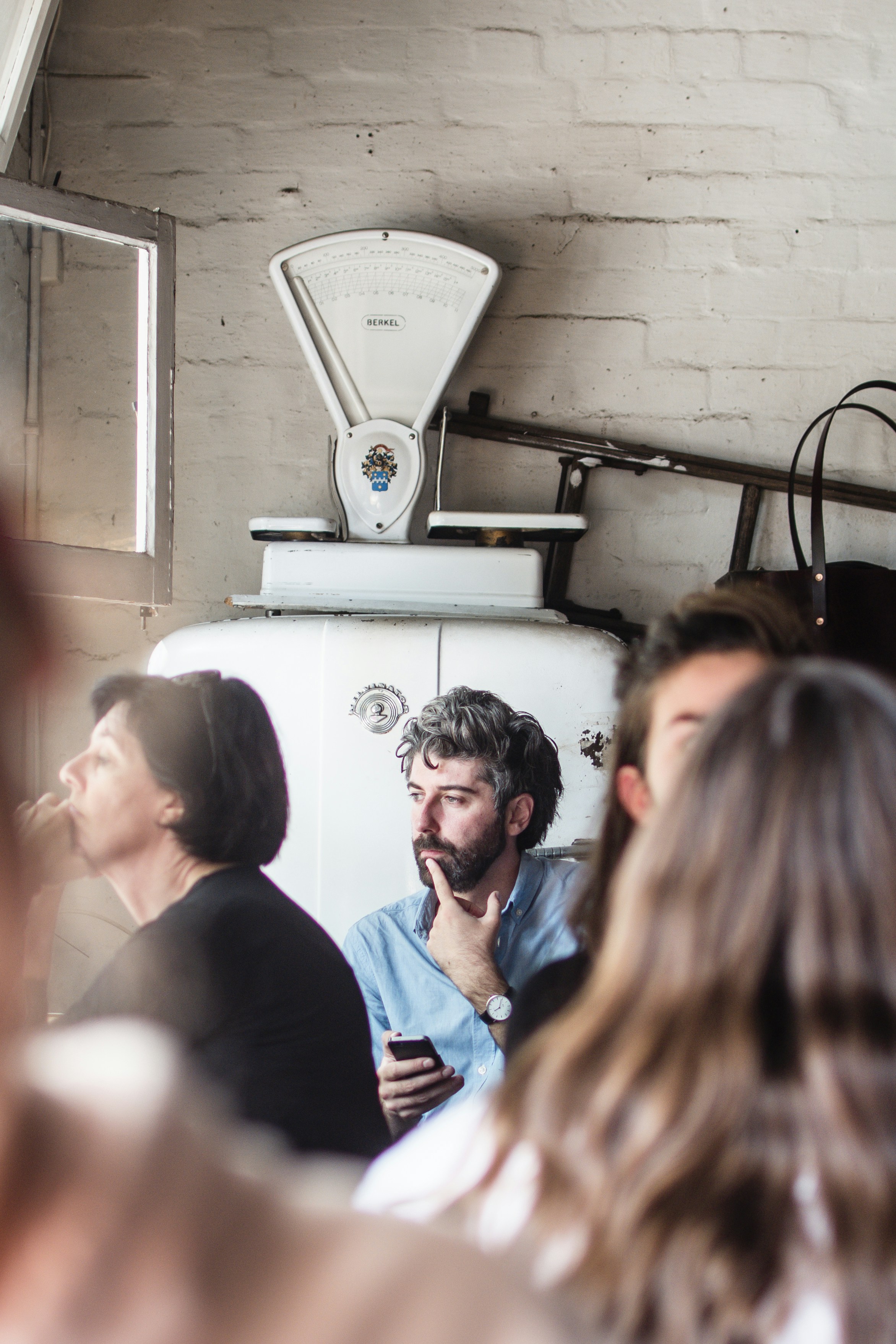 A pensive man in a blue shirt sits amidst a bustling café scene, with an old scale and vintage decor in the background.