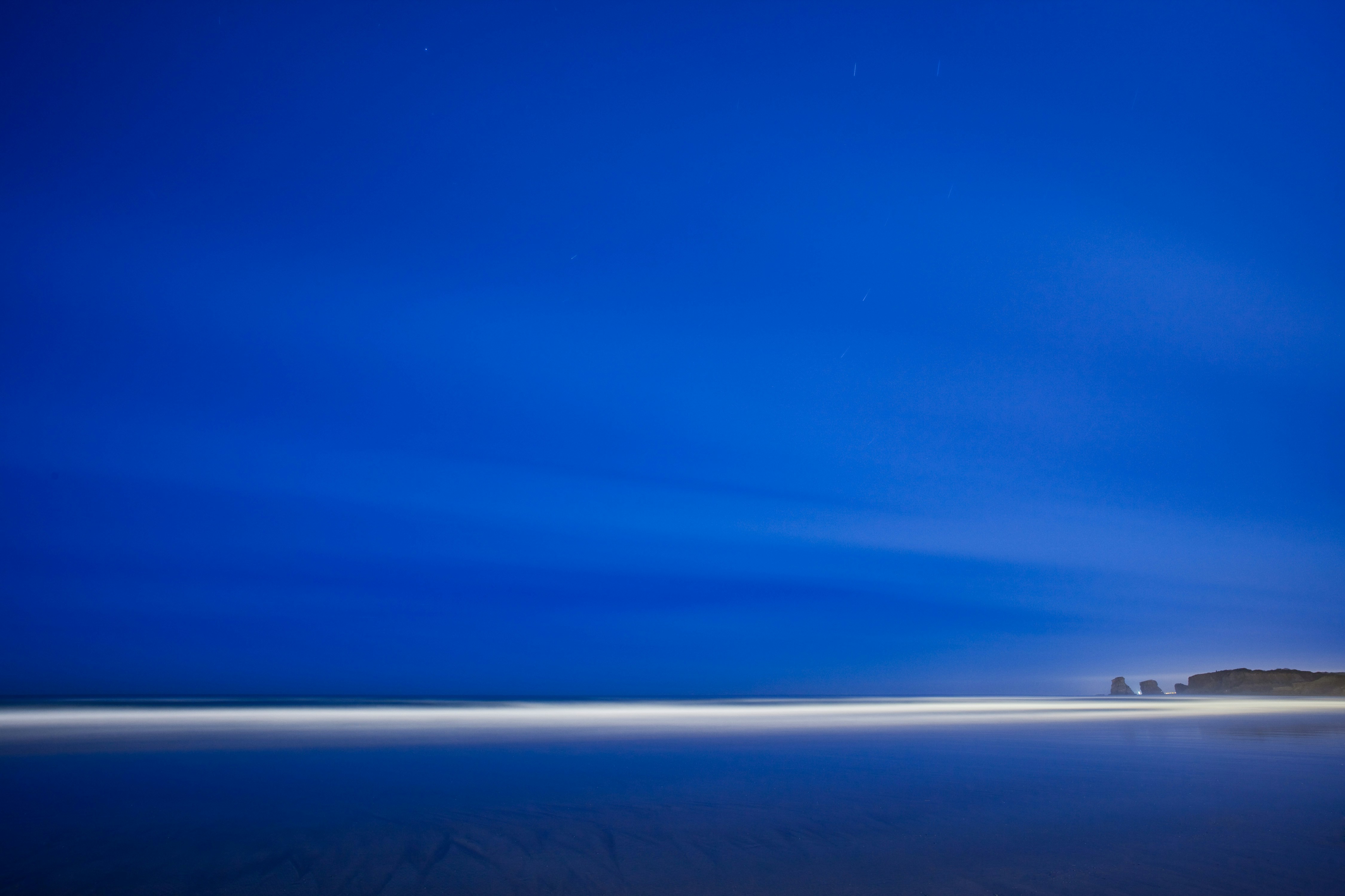 Gentle waves lap against the shore under a deep blue twilight sky, with distant silhouettes of rocks peeking through the haze.