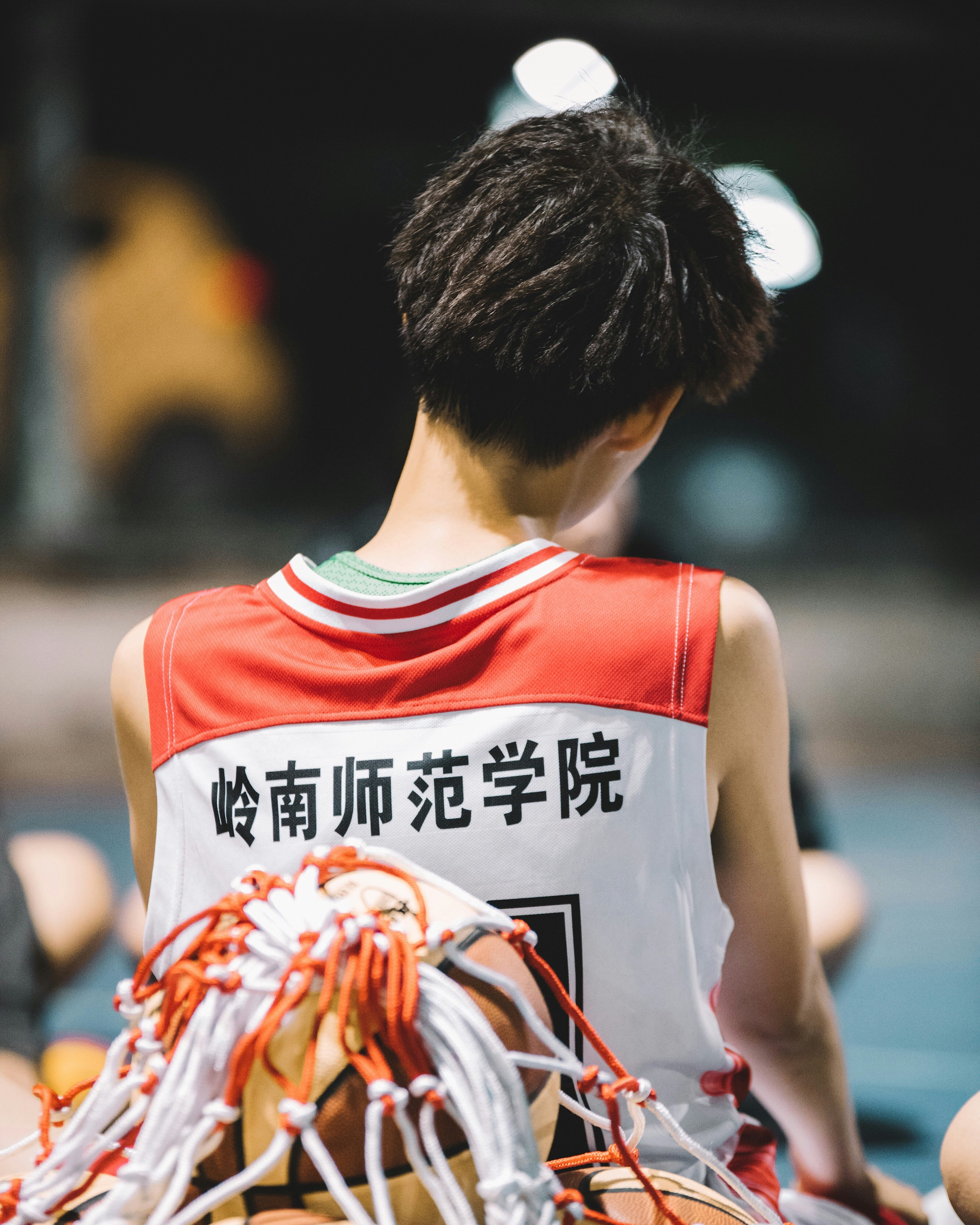 A young basketball player sits with their back to the camera, wearing a jersey that displays their school's name, while a basketball net rests nearby.