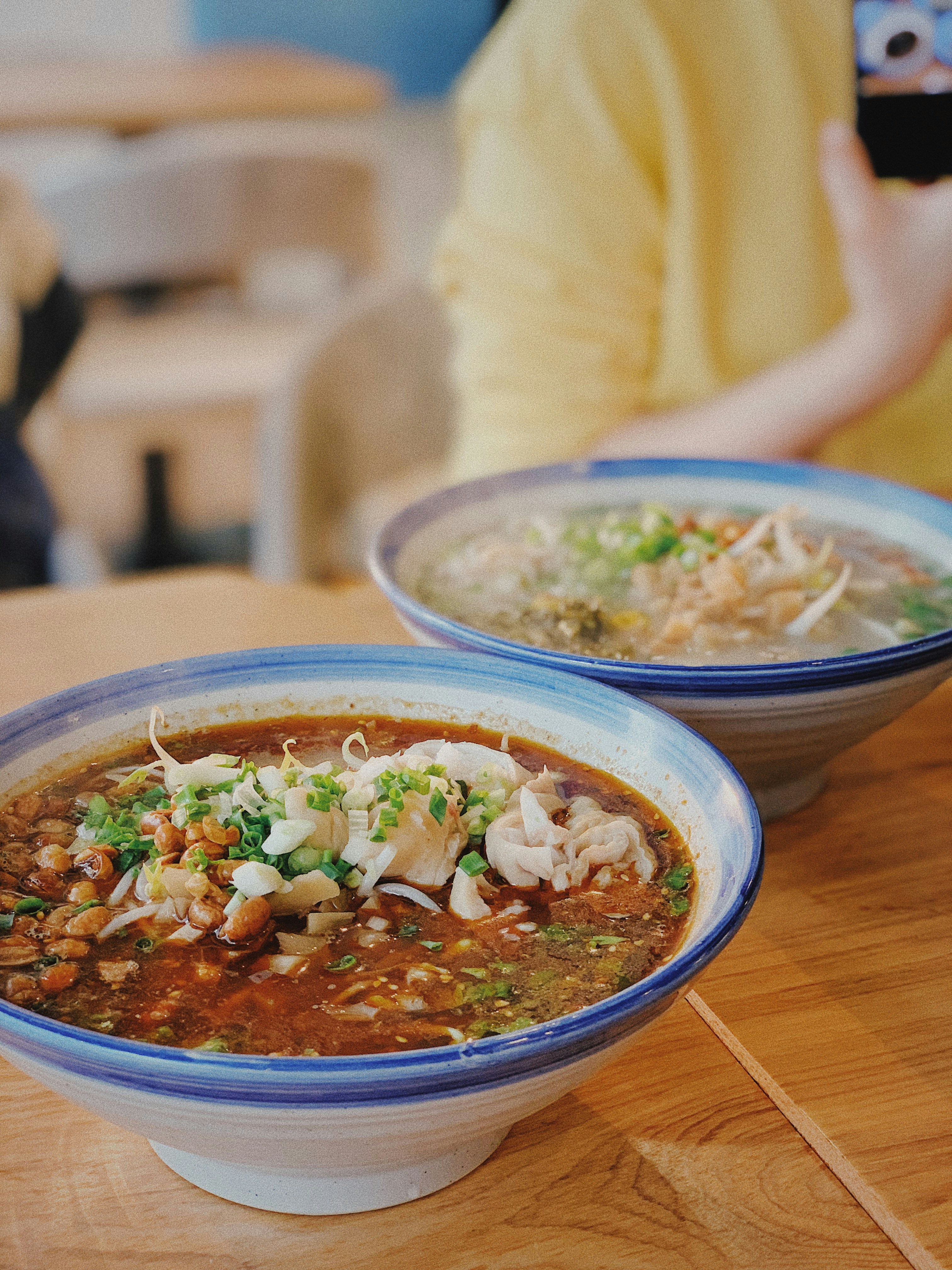 Two bowls of vibrant noodle dishes sit on a wooden table, showcasing a mix of ingredients and textures. A hand with a camera captures the moment in the background.