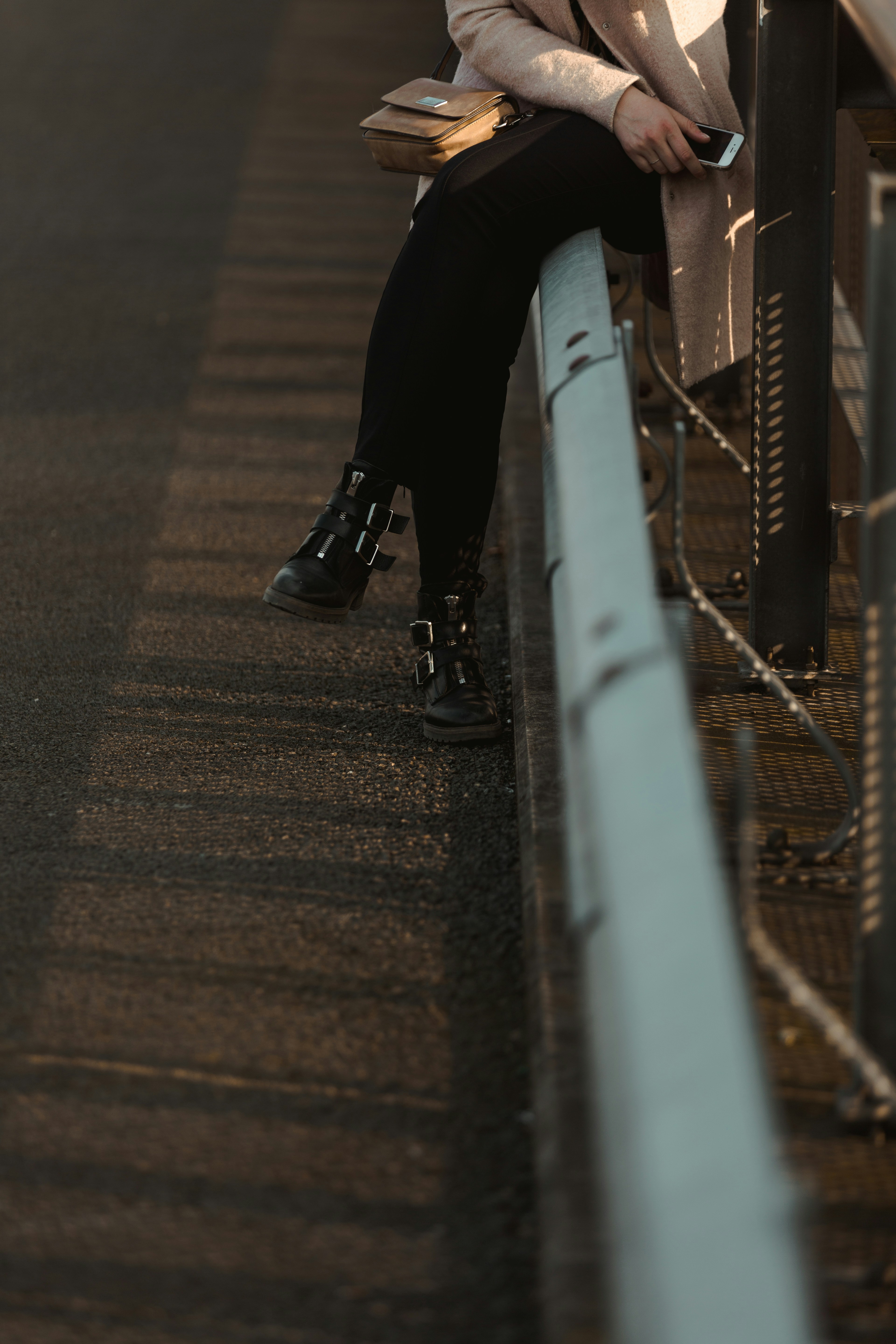 A person sitting on a railing, showcasing stylish black boots and casual attire, with soft evening light casting shadows on the surface.