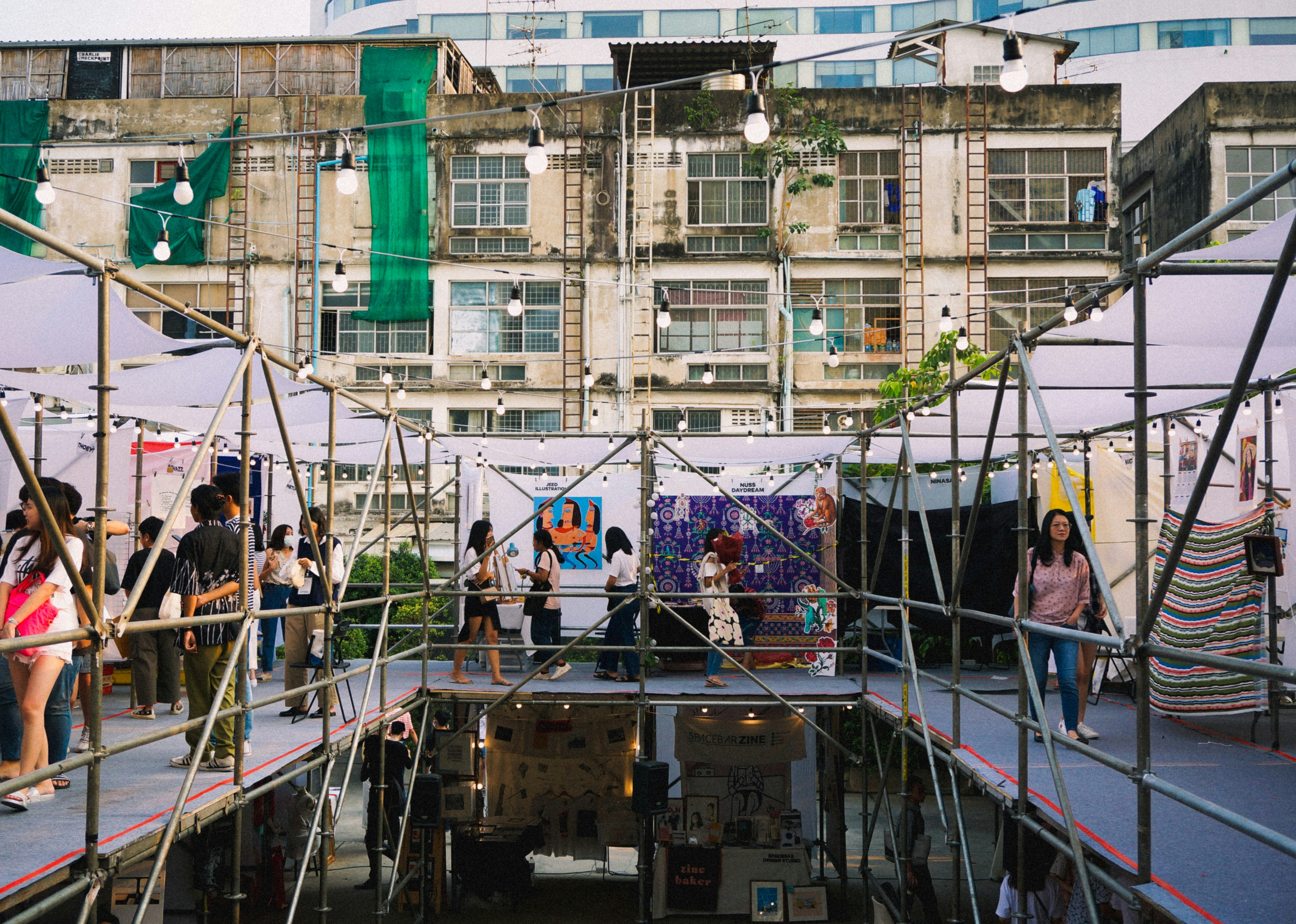 A bustling urban market scene featuring various stalls and visitors exploring artistic displays. The backdrop showcases a mix of modern and vintage architecture.