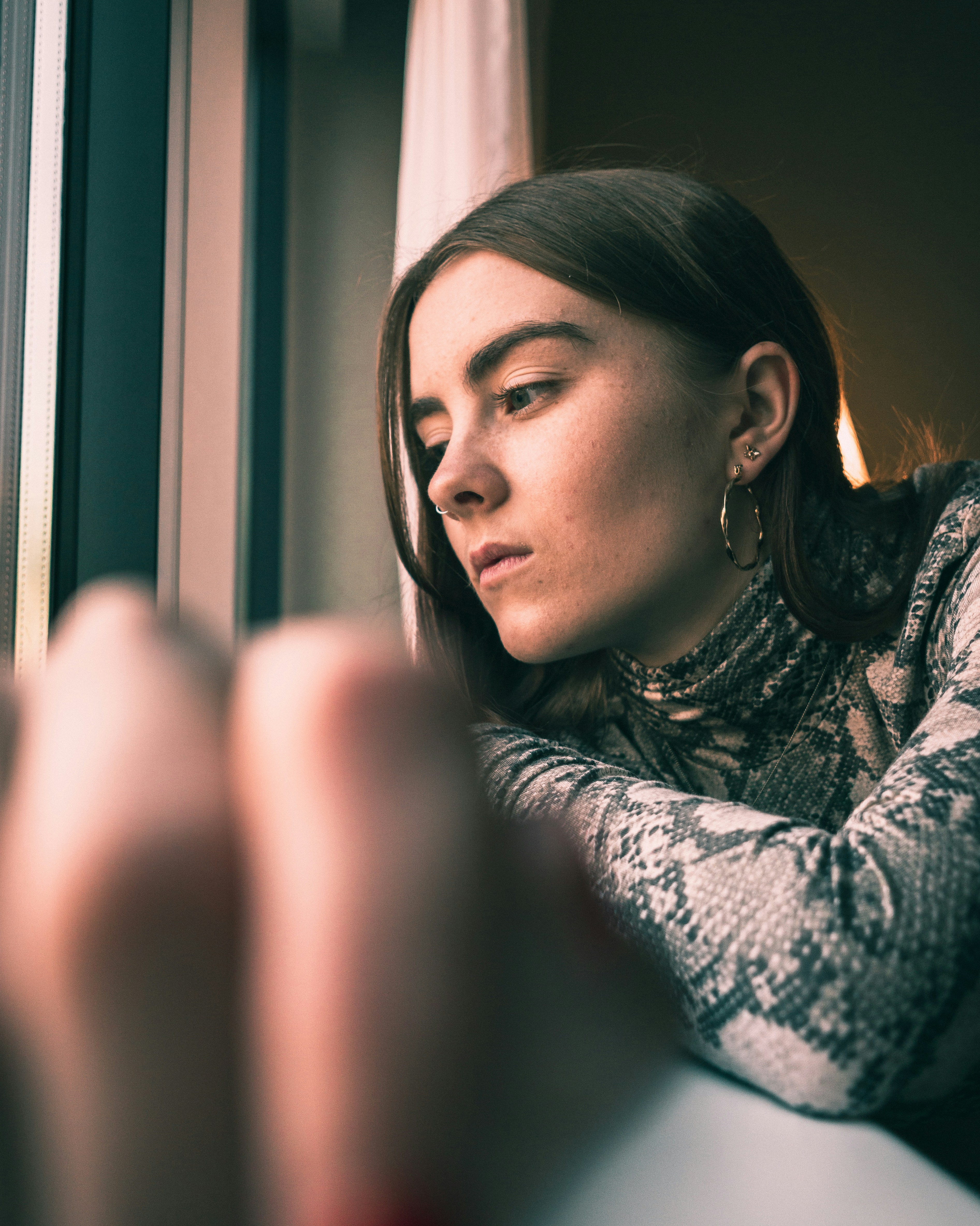 A young woman gazes thoughtfully out the window, her expression reflecting introspection. The scene captures a blend of natural light and subtle details in her attire.
