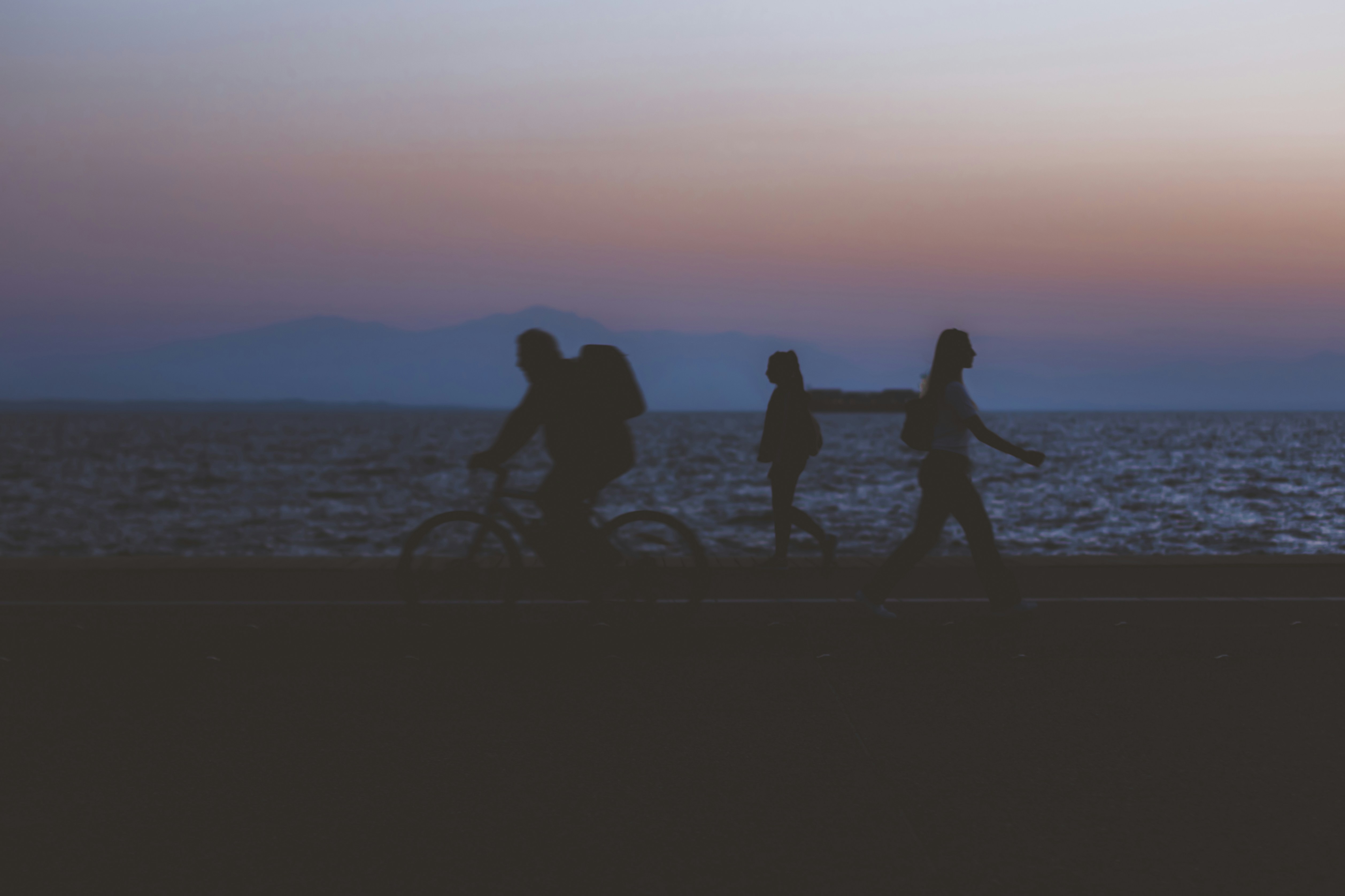 Silhouetted figures walking along a waterfront at dusk, with gentle waves and distant mountains creating a serene backdrop.