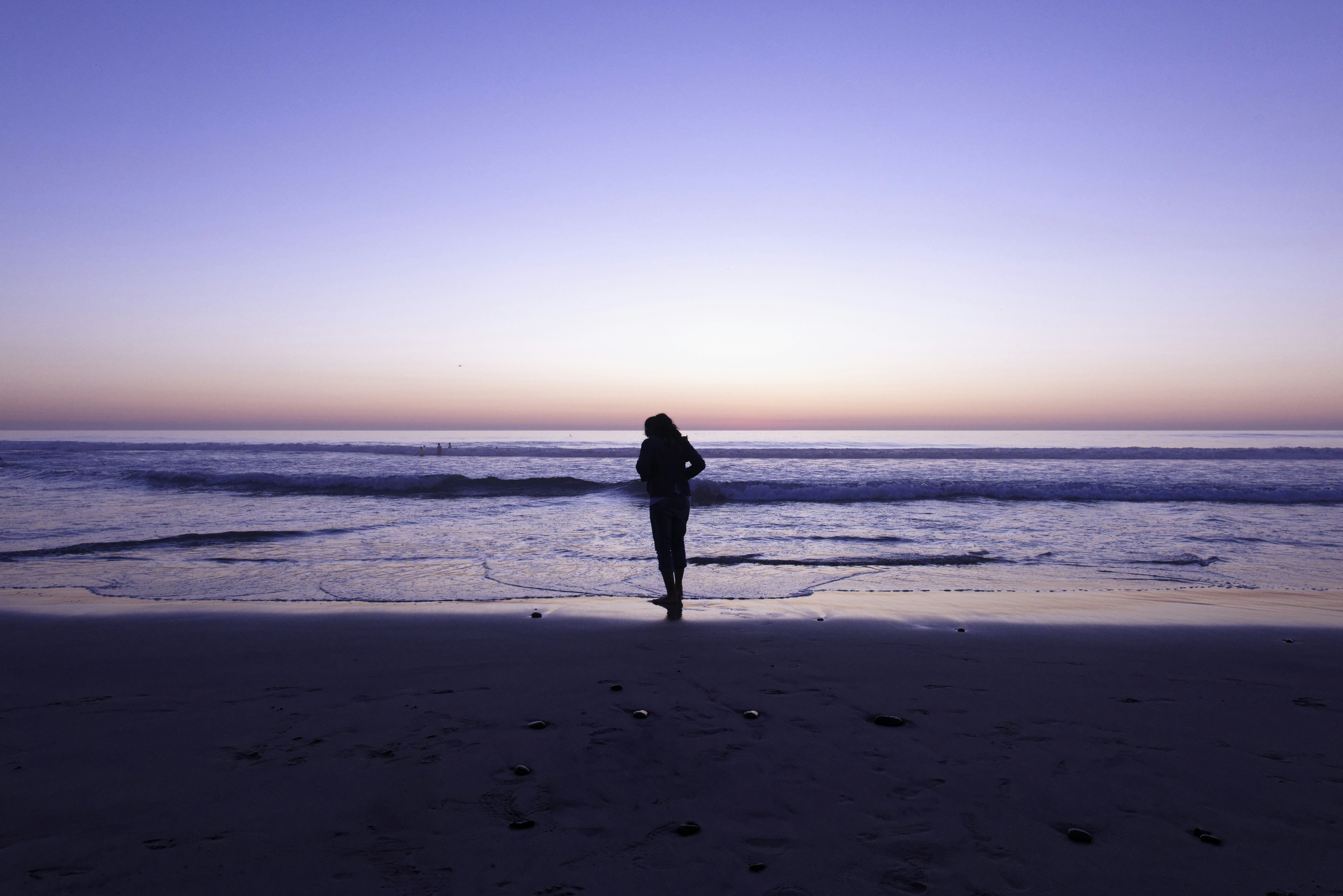 Silhouette of a person standing on a beach during the serene blue hour, with gentle waves and a gradient sky.