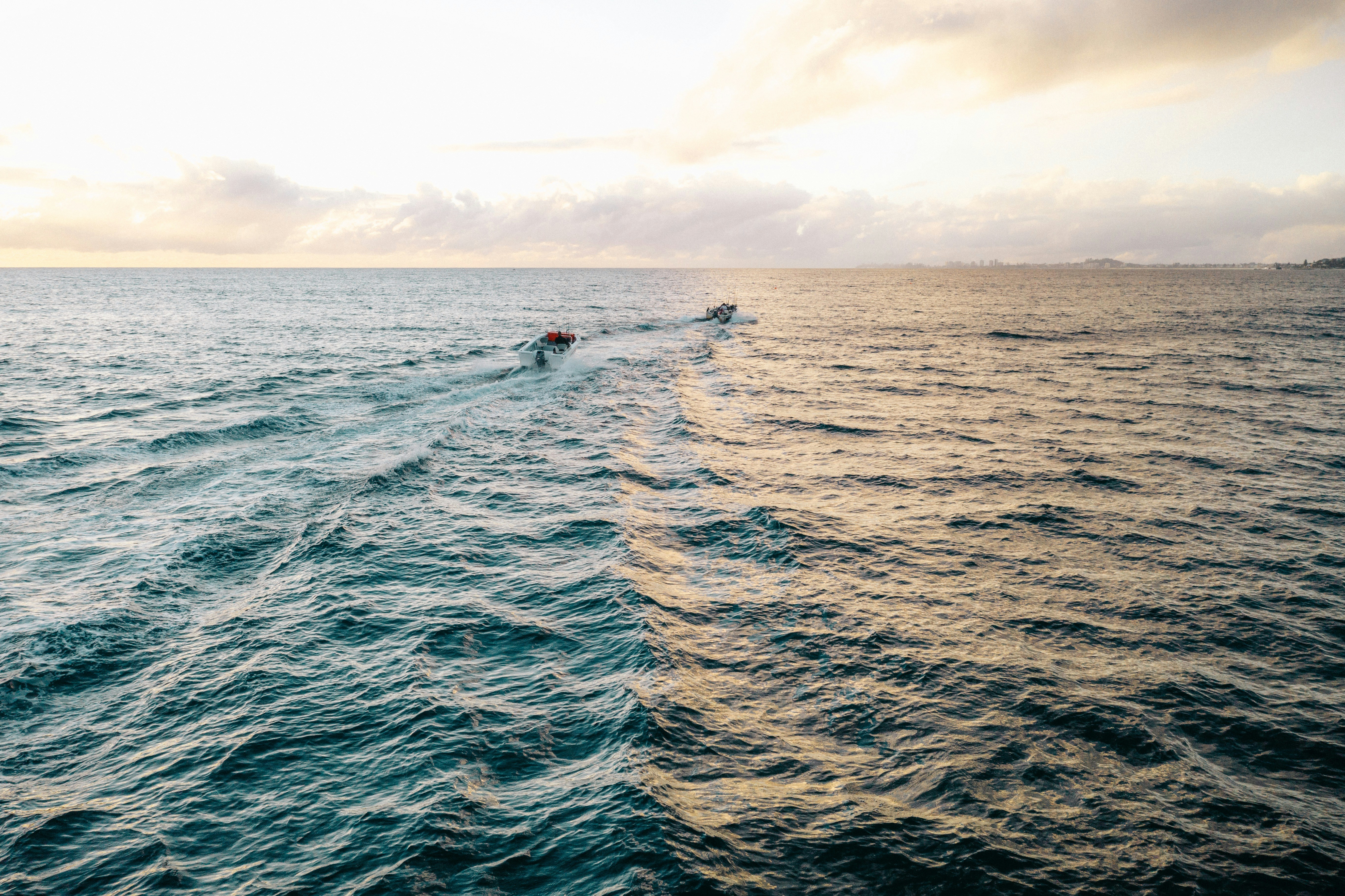 Two boats glide through the tranquil ocean, leaving gentle ripples in their wake as the sun sets on the horizon.