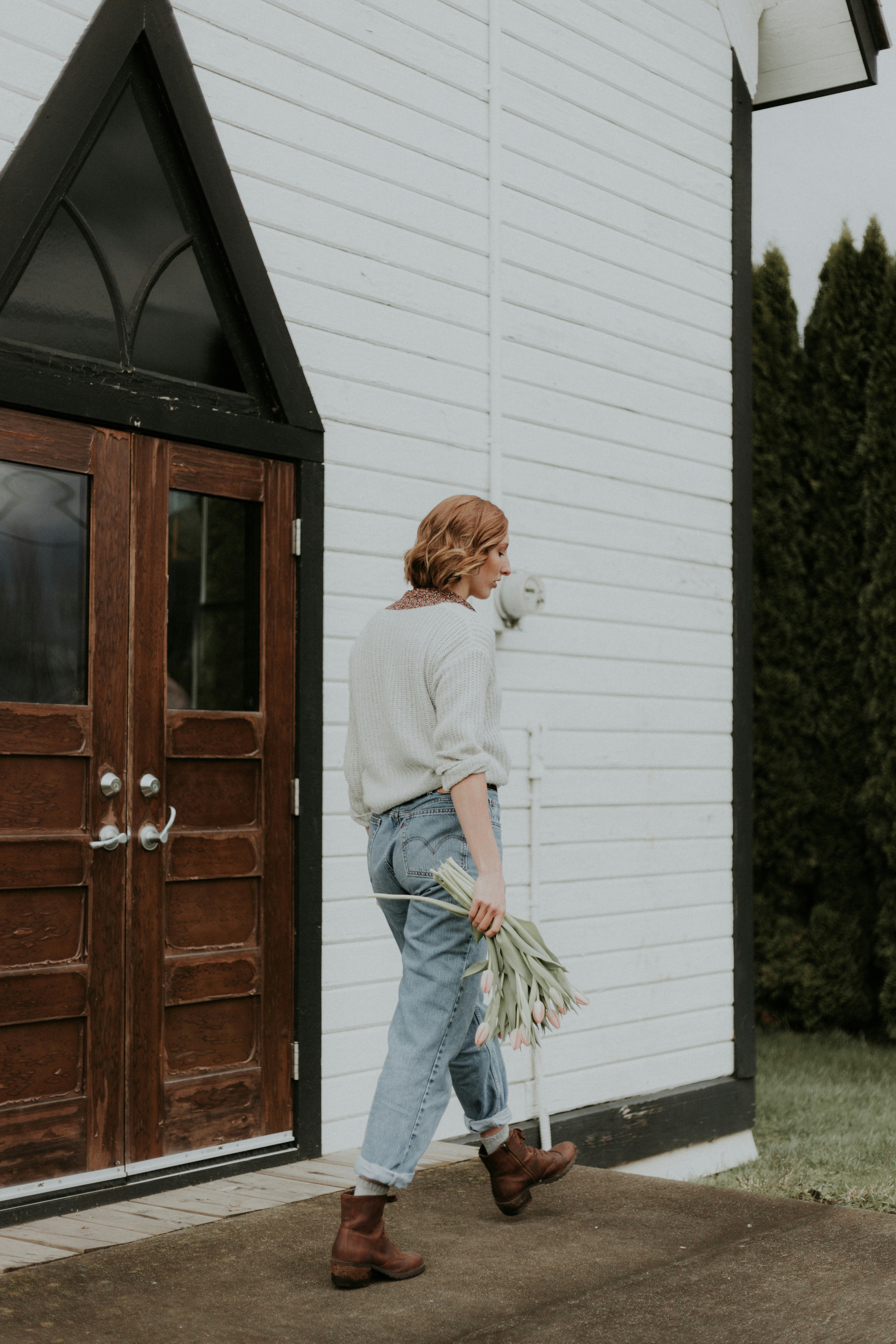 Woman in casual attire carrying fresh flowers while walking past a charming white building with wooden doors.