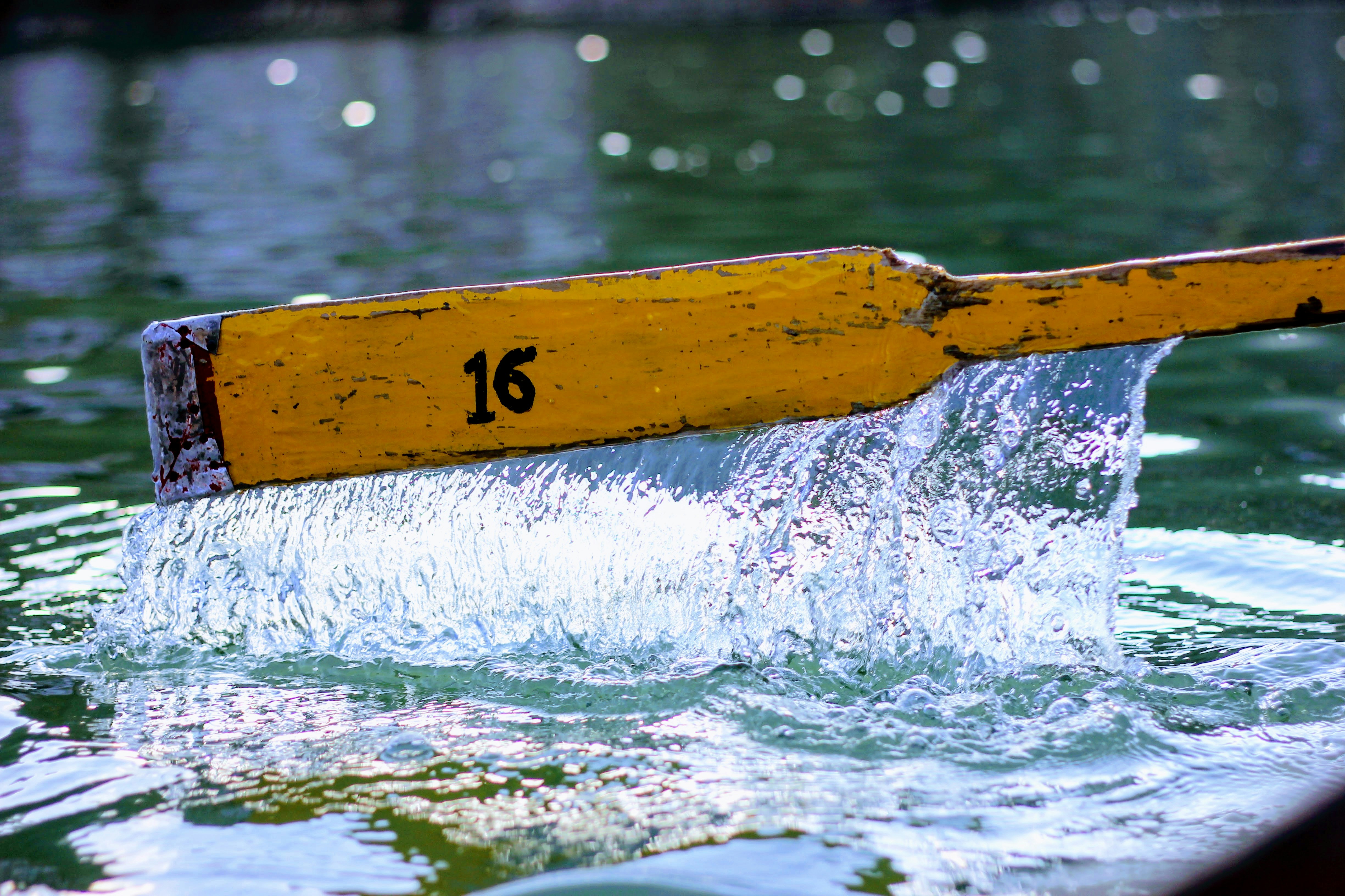 Yellow rowing oar slicing through the water, creating dynamic splashes and ripples under sunlight.