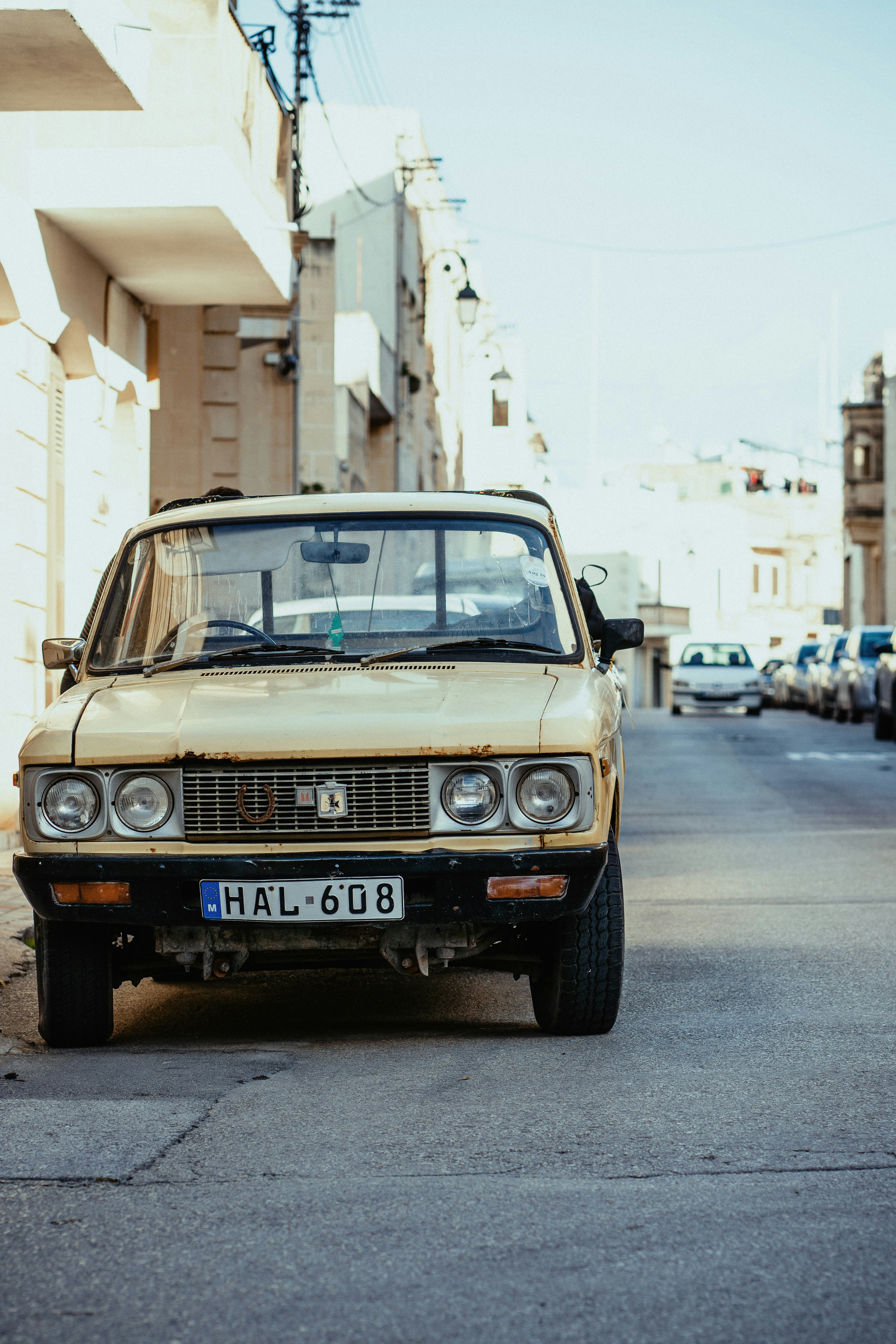 Vintage yellow car parked on a narrow street, surrounded by charming architecture and other vehicles in the background.